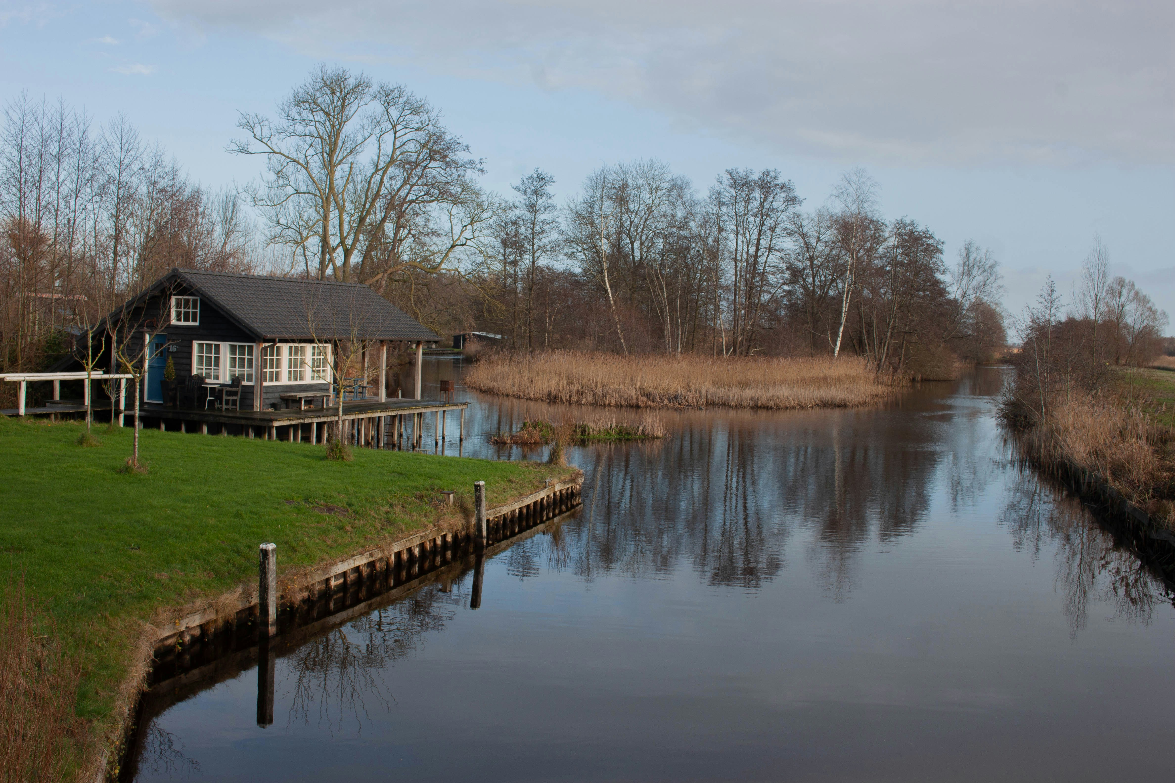 A serene cabin nestled by a calm river, surrounded by leafless trees under a partly cloudy sky.