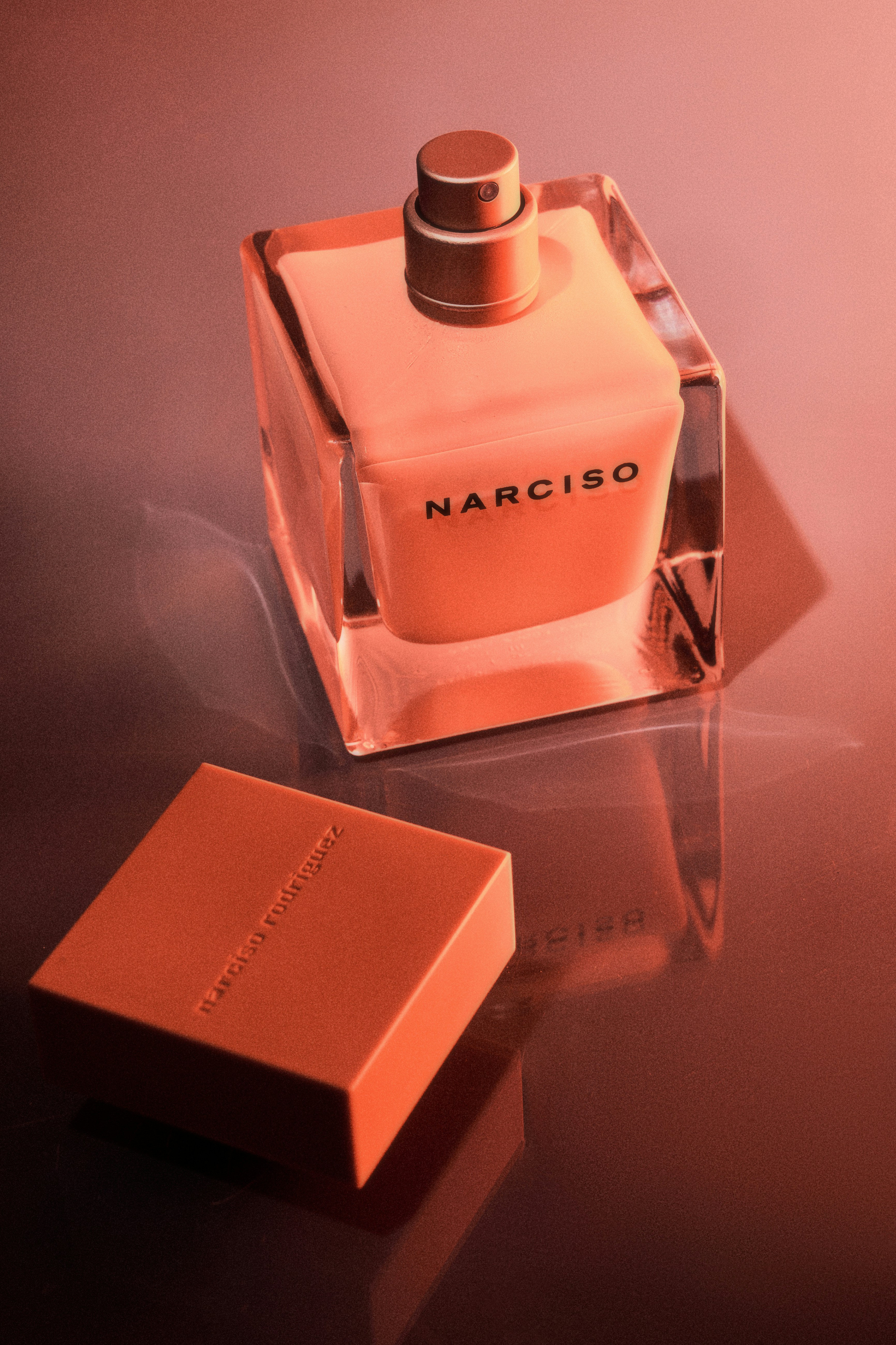 Photograph of a Narciso perfume bottle and a matching box resting on a reflective surface under magenta lighting.