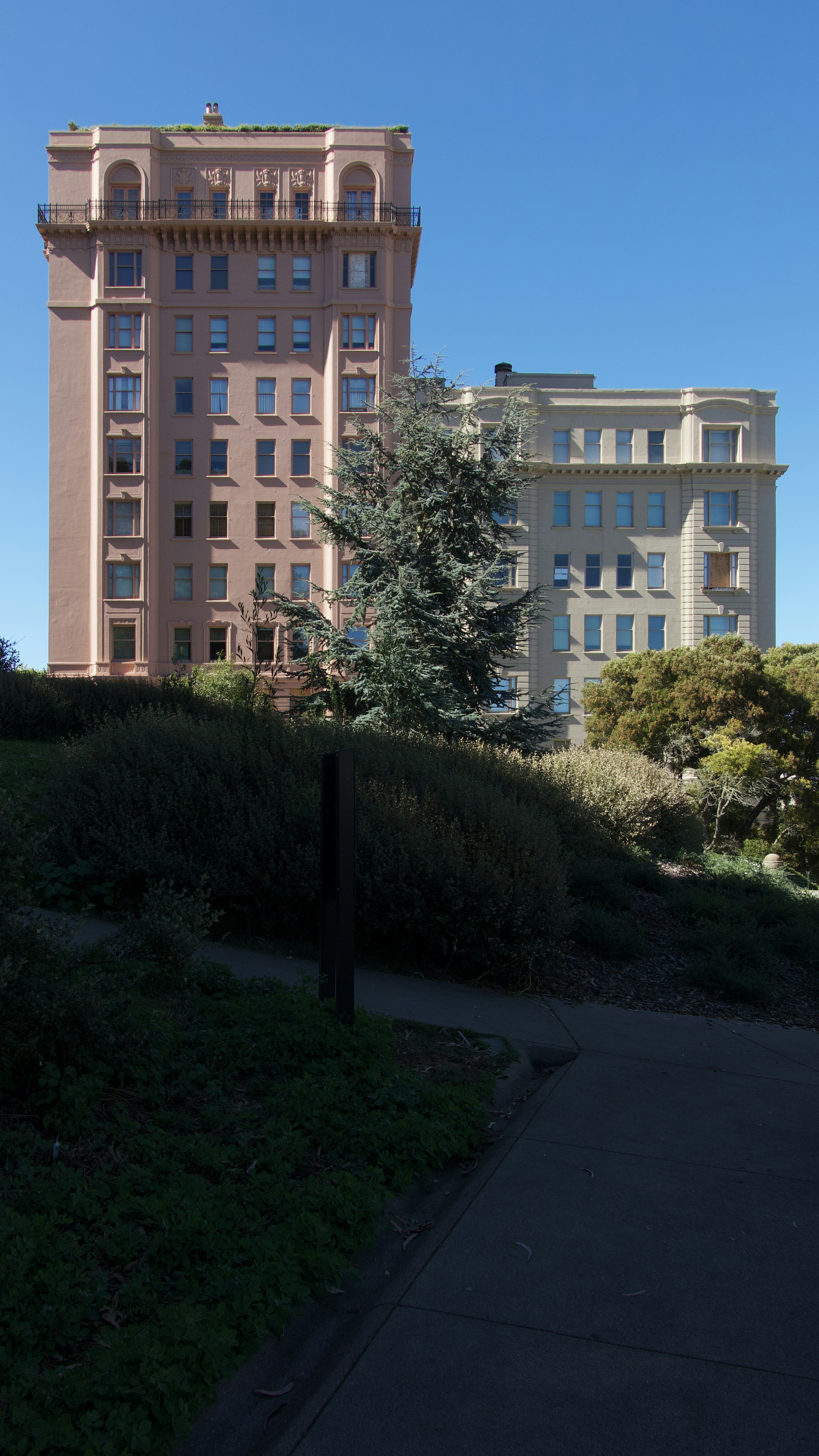 Tall pink and beige buildings rise behind a lush urban park under a clear blue sky.