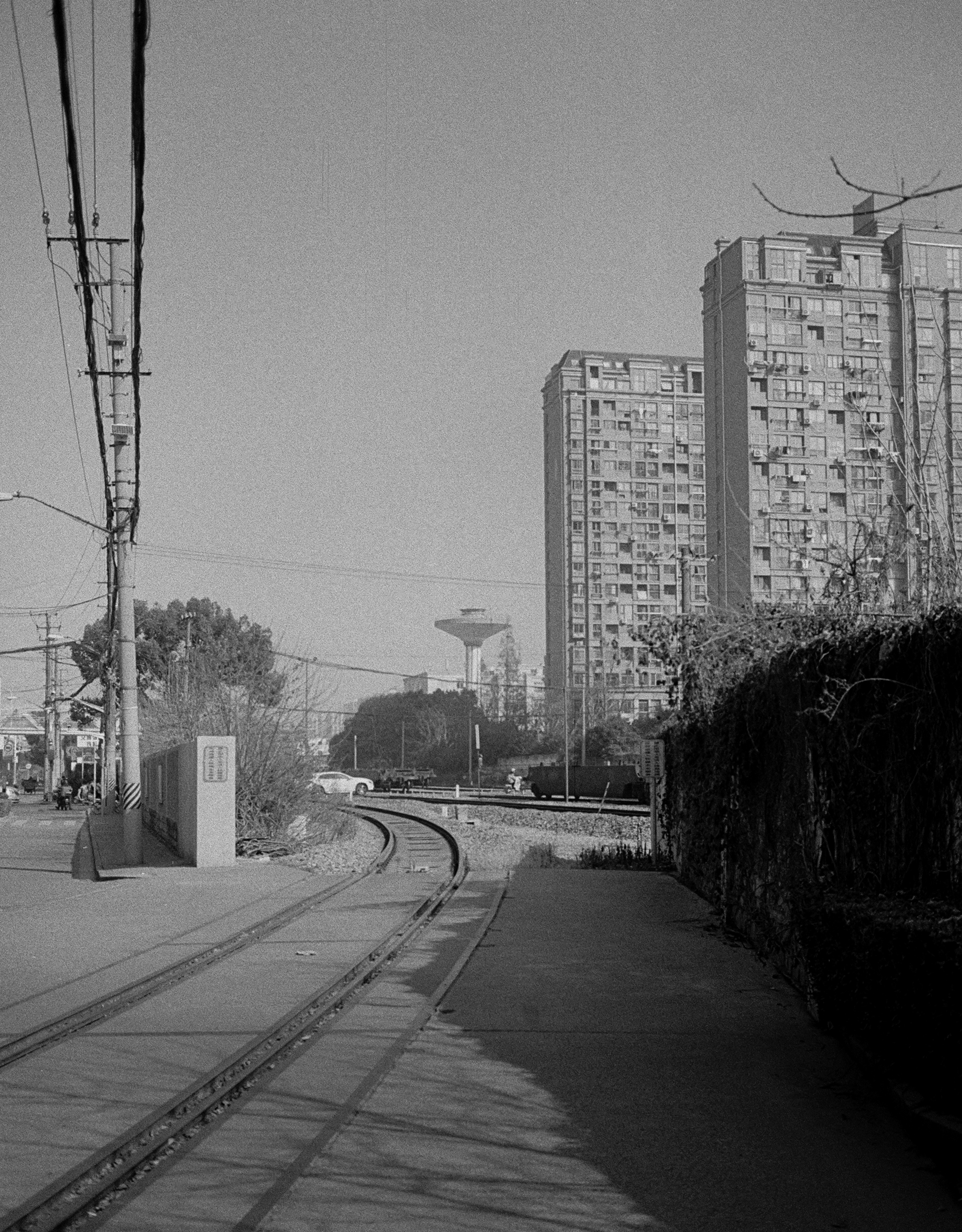 Monochrome street scene with tram tracks curving toward distant apartment blocks. A hedge and overhead lines frame the composition.