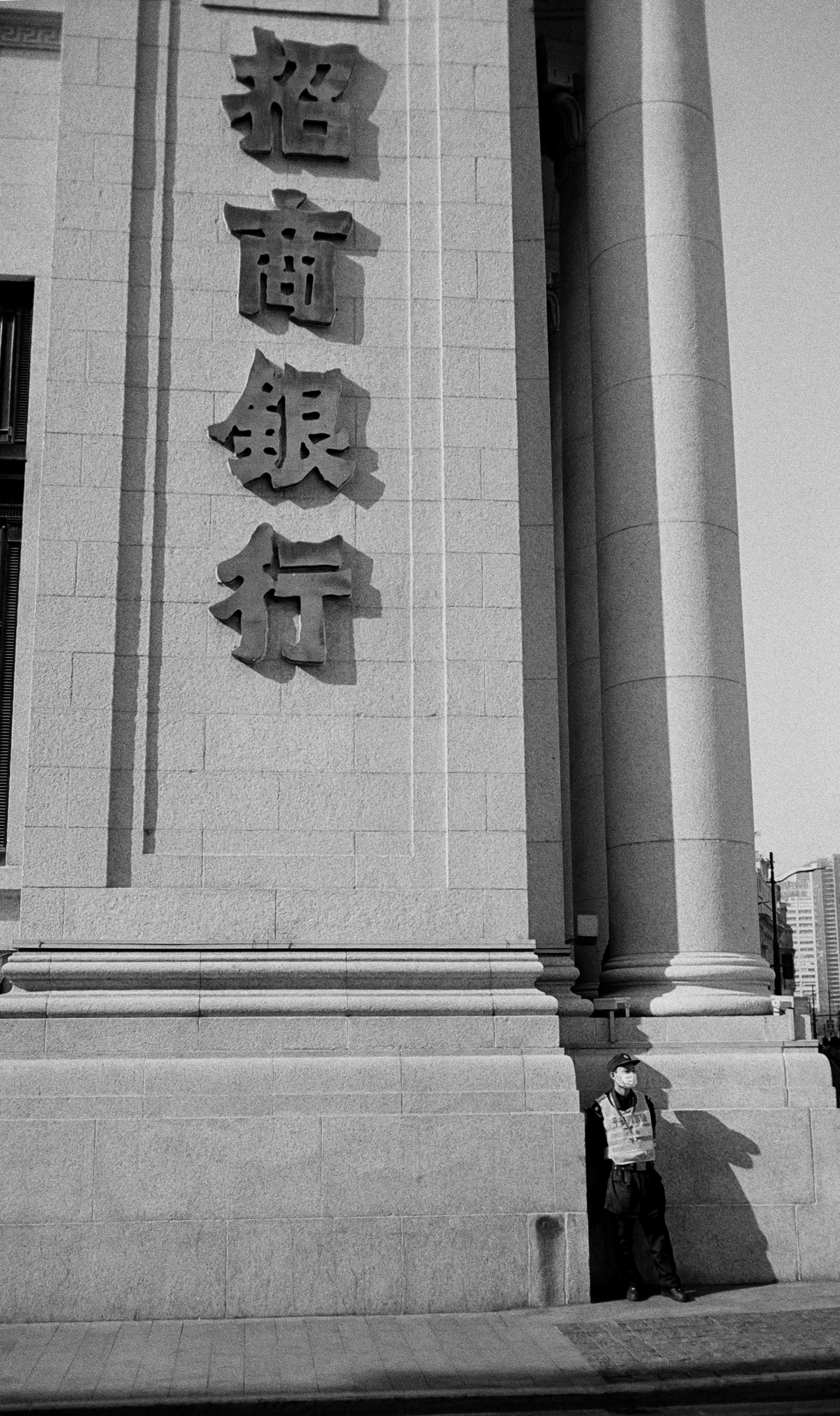 Black-and-white photograph of a security guard leaning against the stone base of a monumental bank facade, with tall Chinese characters carved into the wall. The scene emphasizes scale and quiet vigilance.