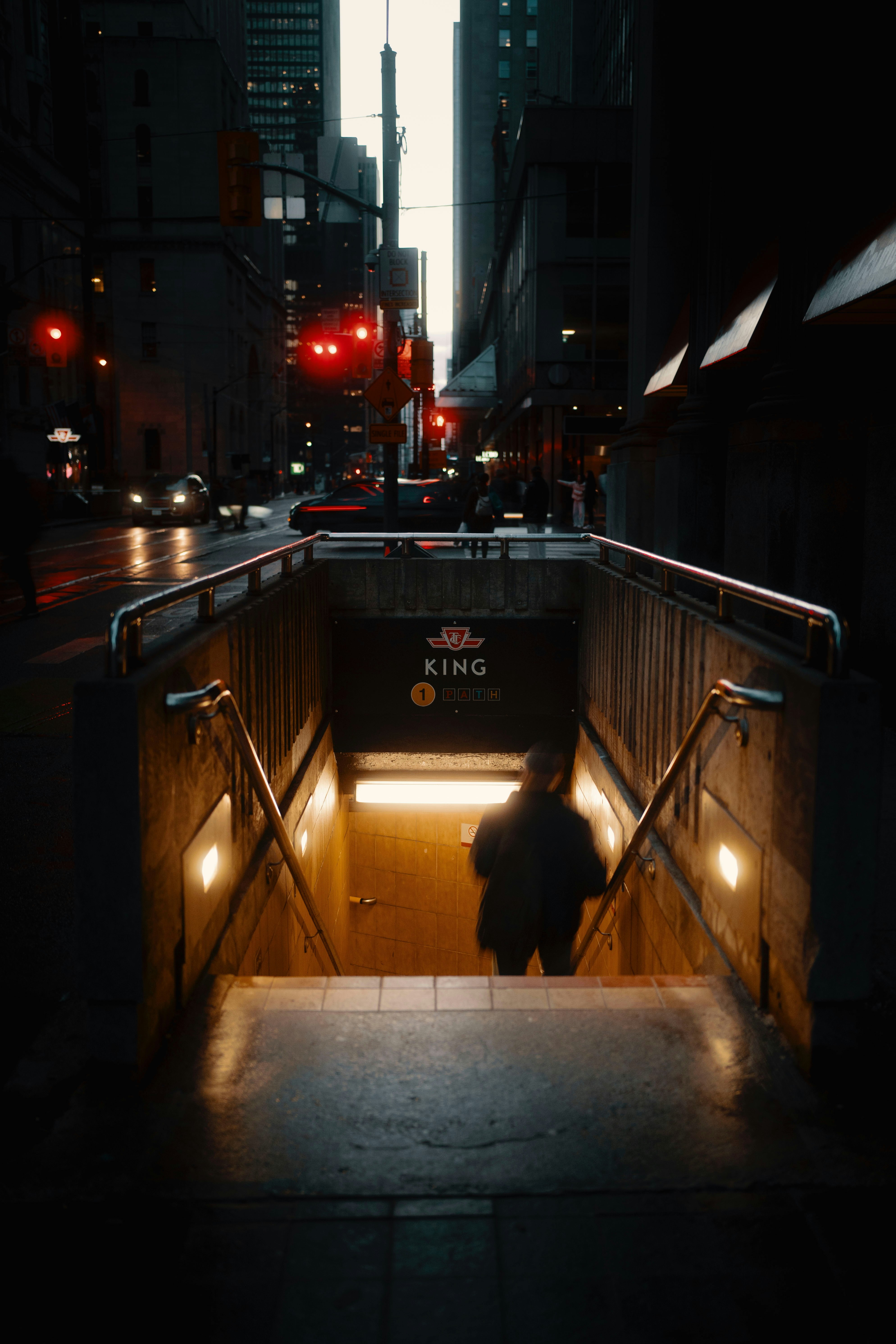 Lone figure descending into a dimly lit subway entrance, framed by a bustling cityscape at dusk. Warm underground lights contrast with cool city tones, enhancing urban energy.