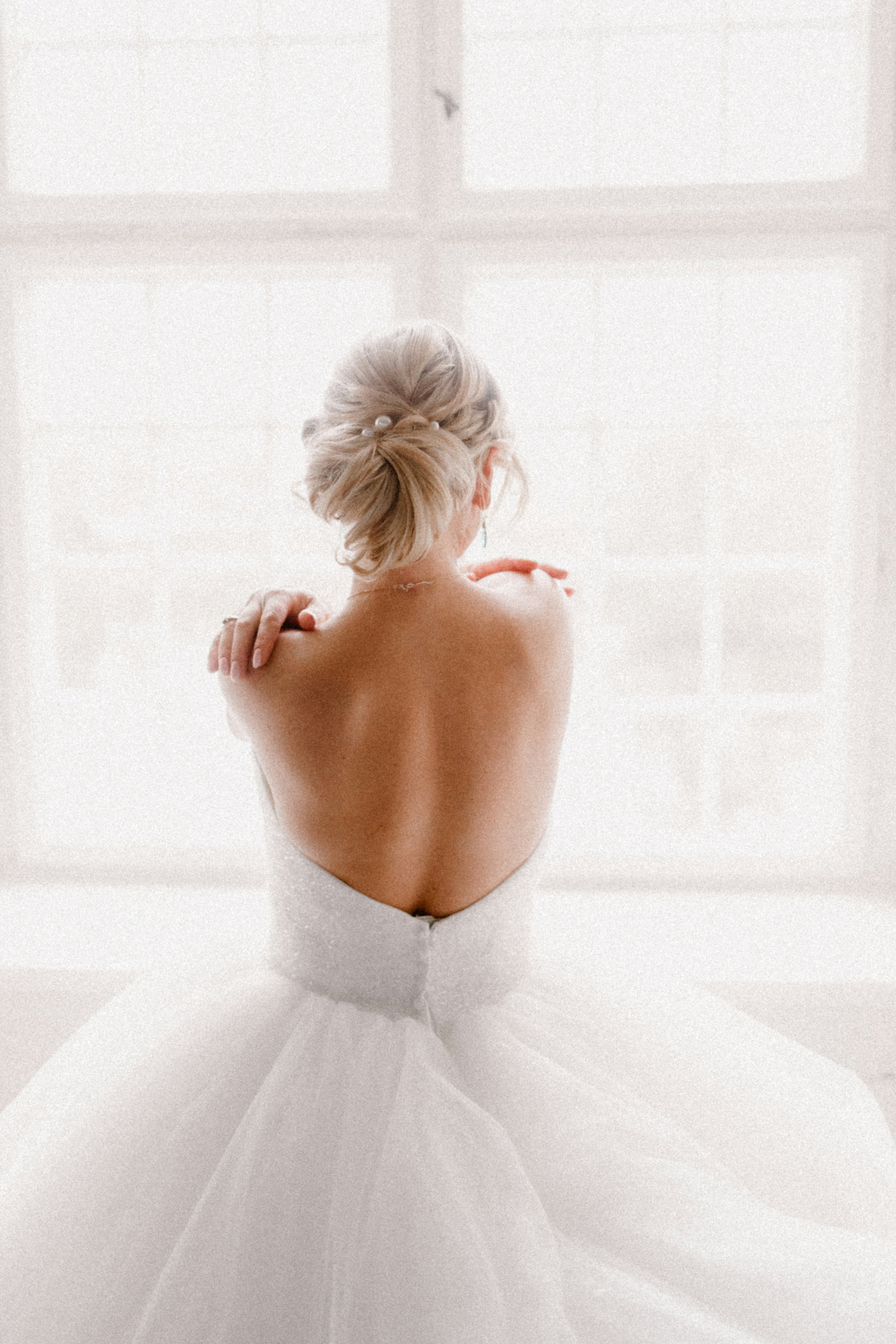Bride in a white gown stands facing a softly lit window, highlighting her elegant hairstyle and the gown's intricacies.