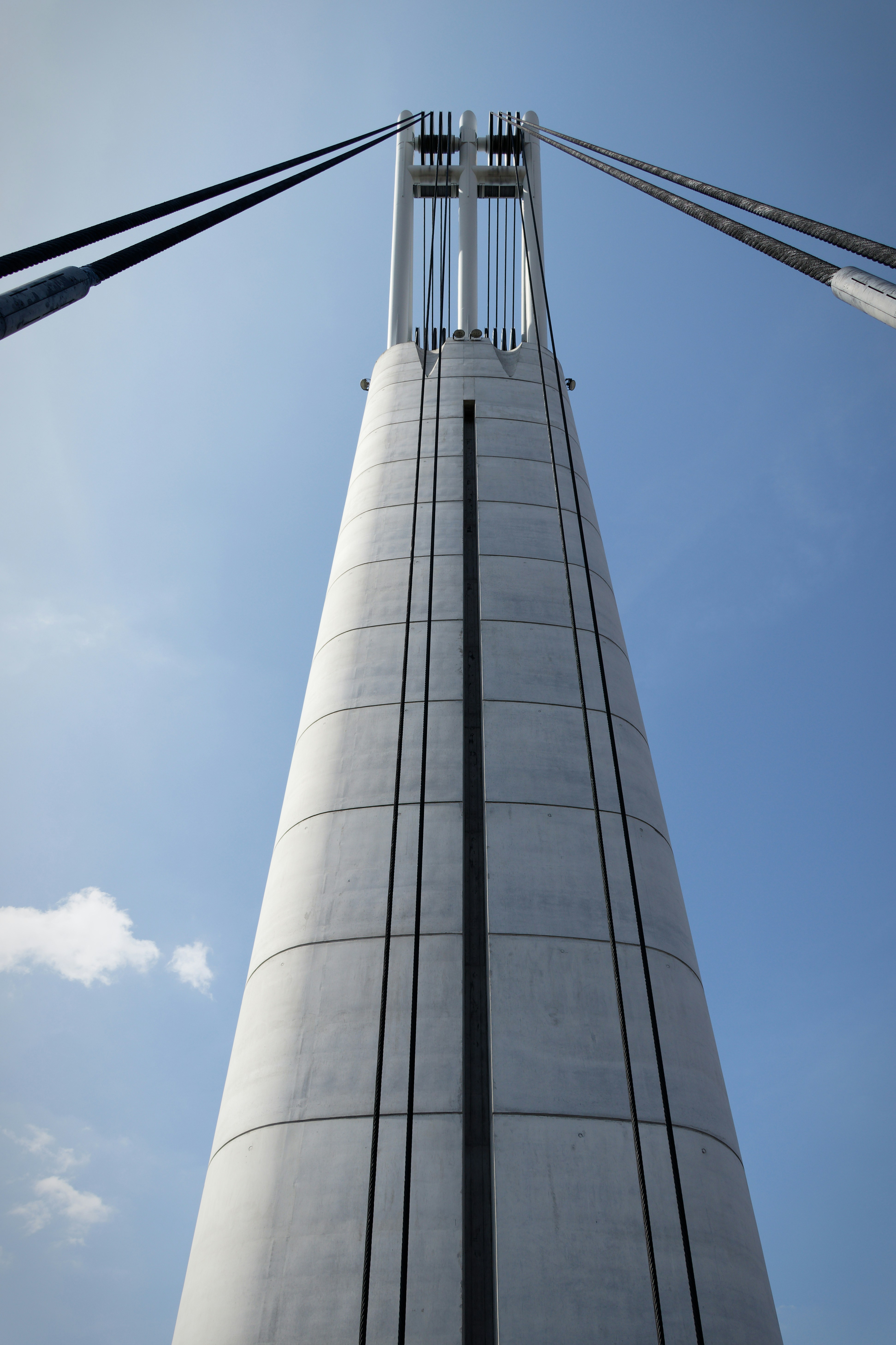Upward view of a massive concrete bridge pillar against a clear blue sky, accented by supporting cables.