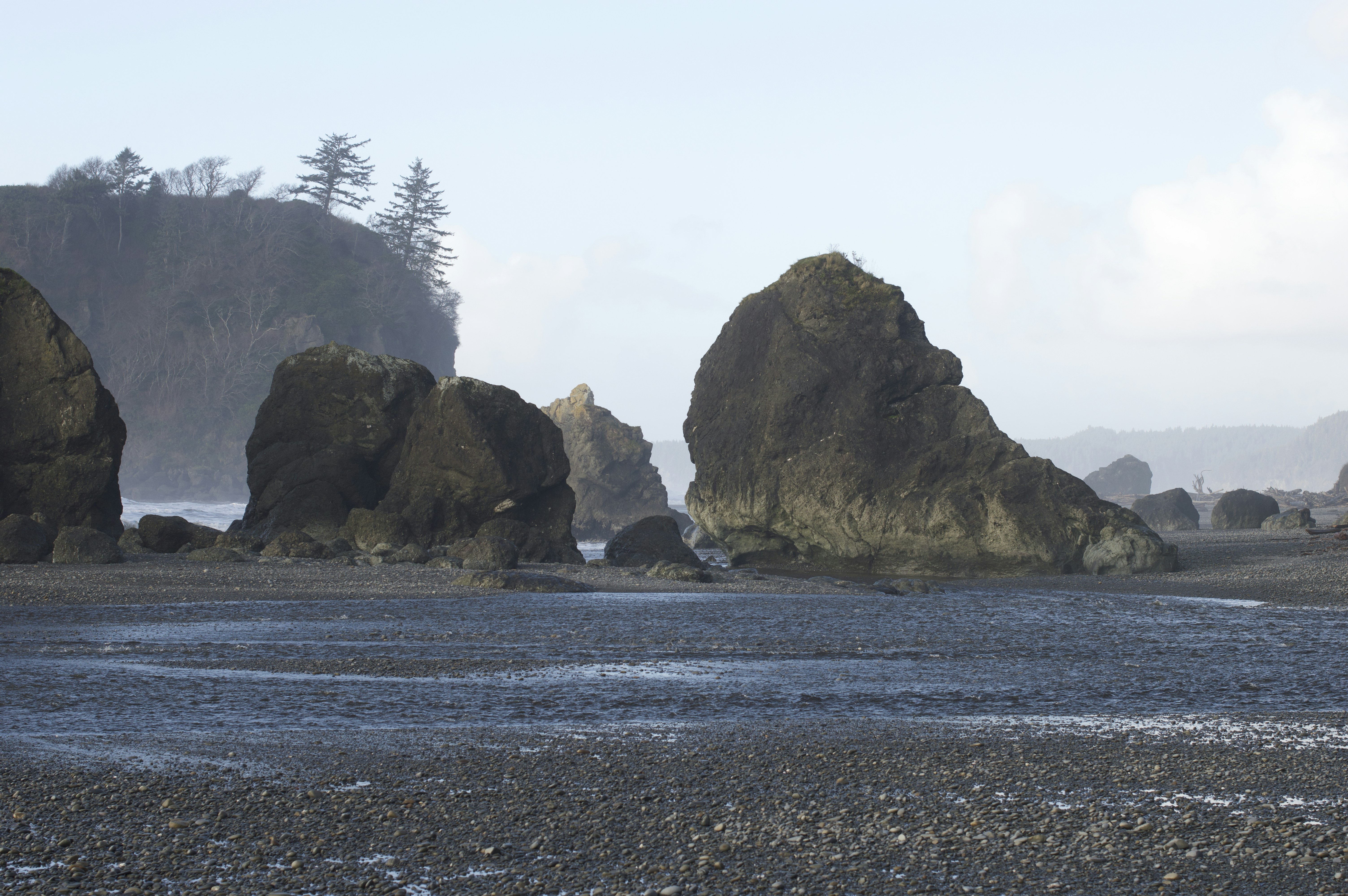 Large rocks scattered along a tranquil beach with distant cliffs and a clear sky.