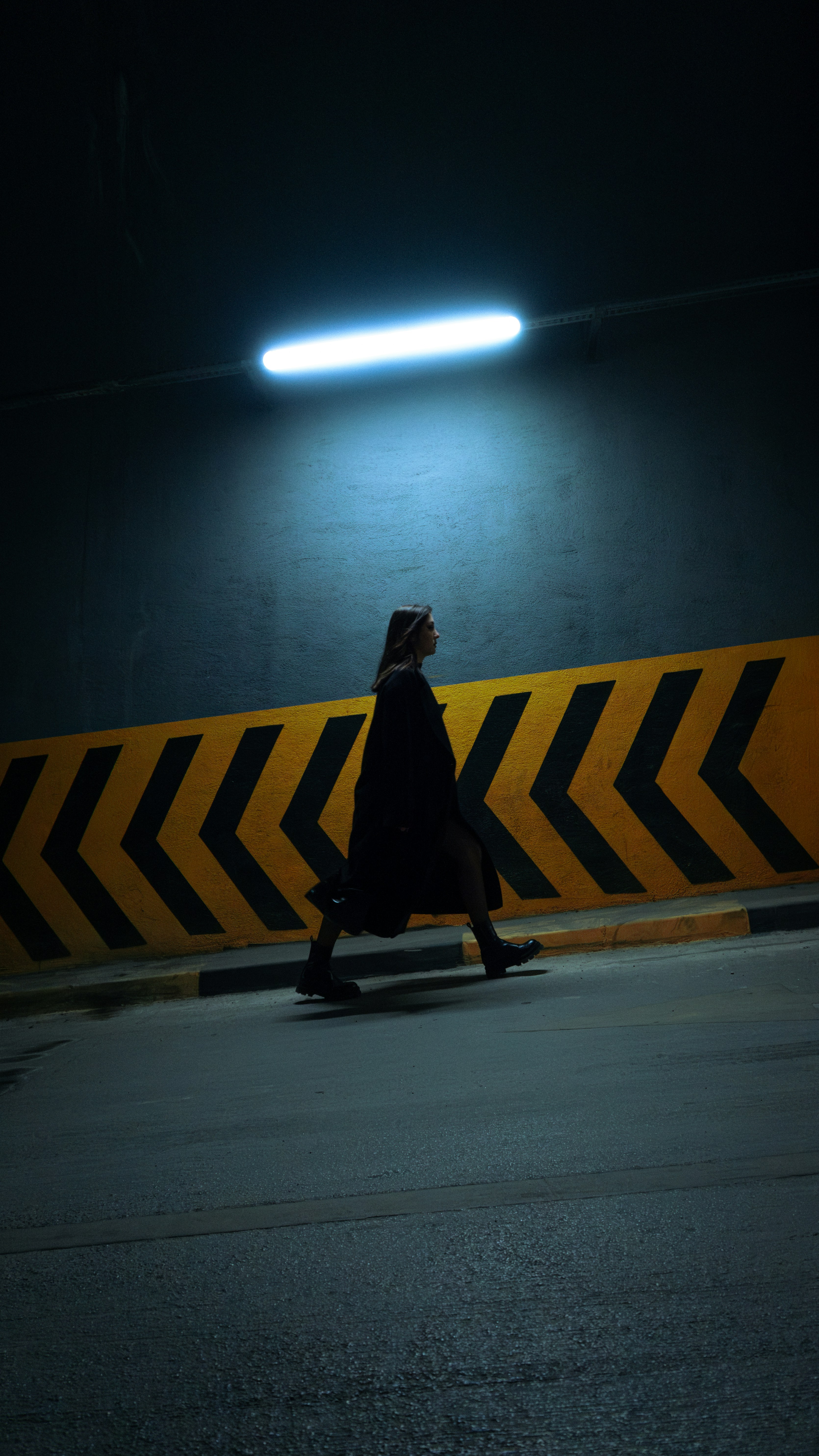Figure walking through a dimly lit urban passage with bold yellow chevrons and a glowing overhead light, creating a cinematic atmosphere.
