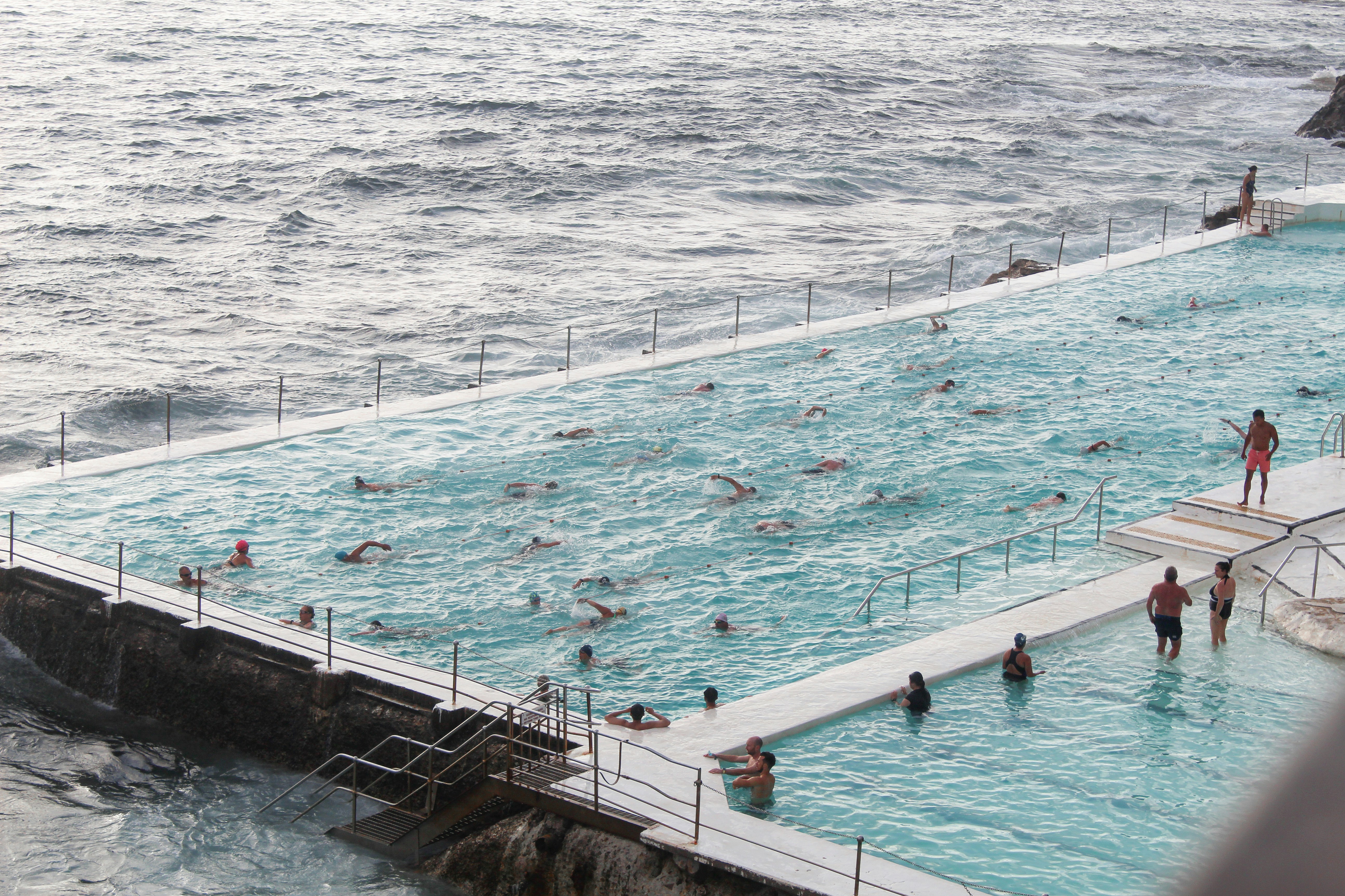 Terraced cliffside pool overlooking the open sea, crowded with swimmers and a few perched on the edge. The composition highlights leisure against rugged coastal architecture.