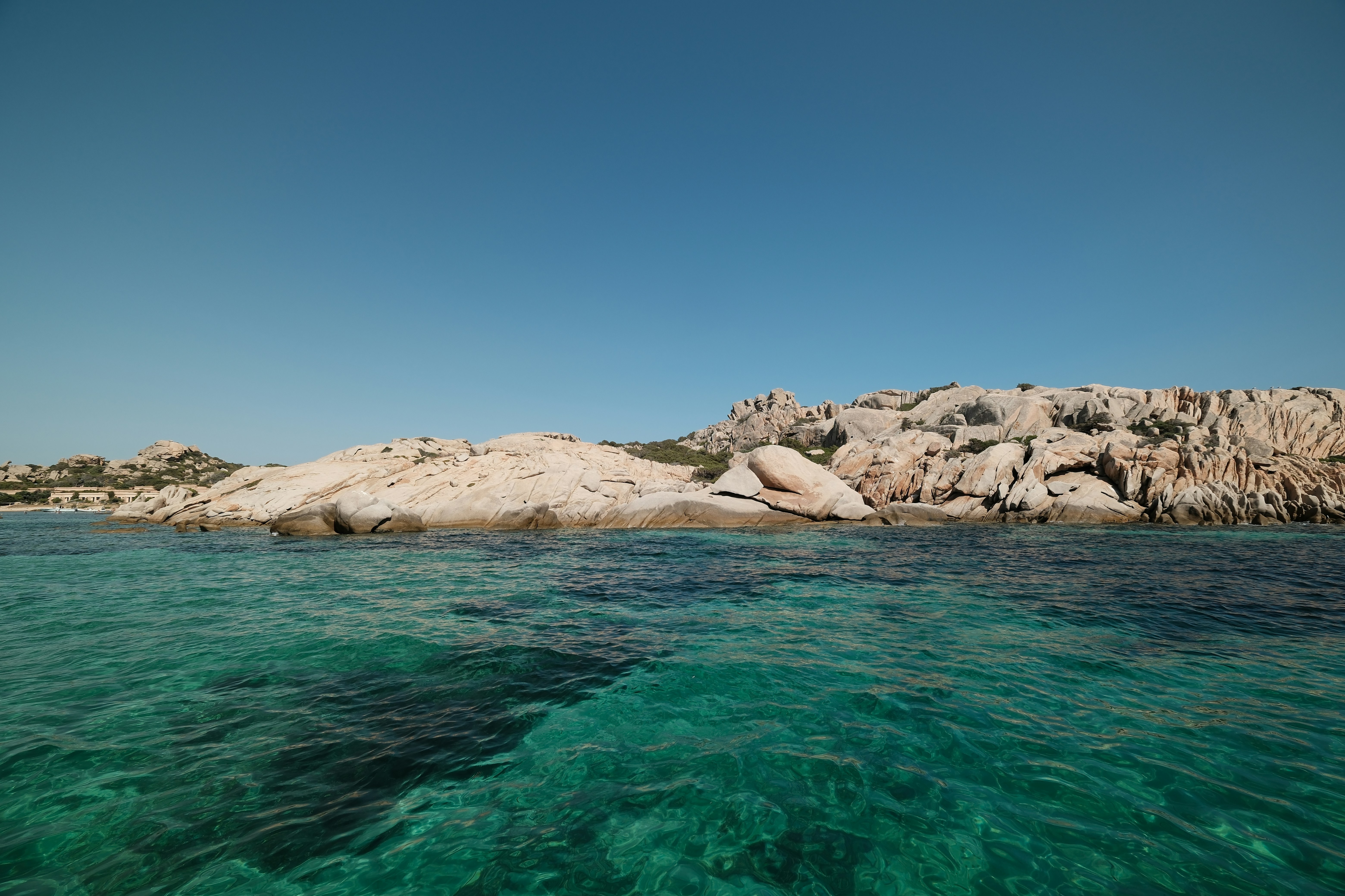 Clear turquoise waters meet rugged rocky coastline under a bright blue sky.