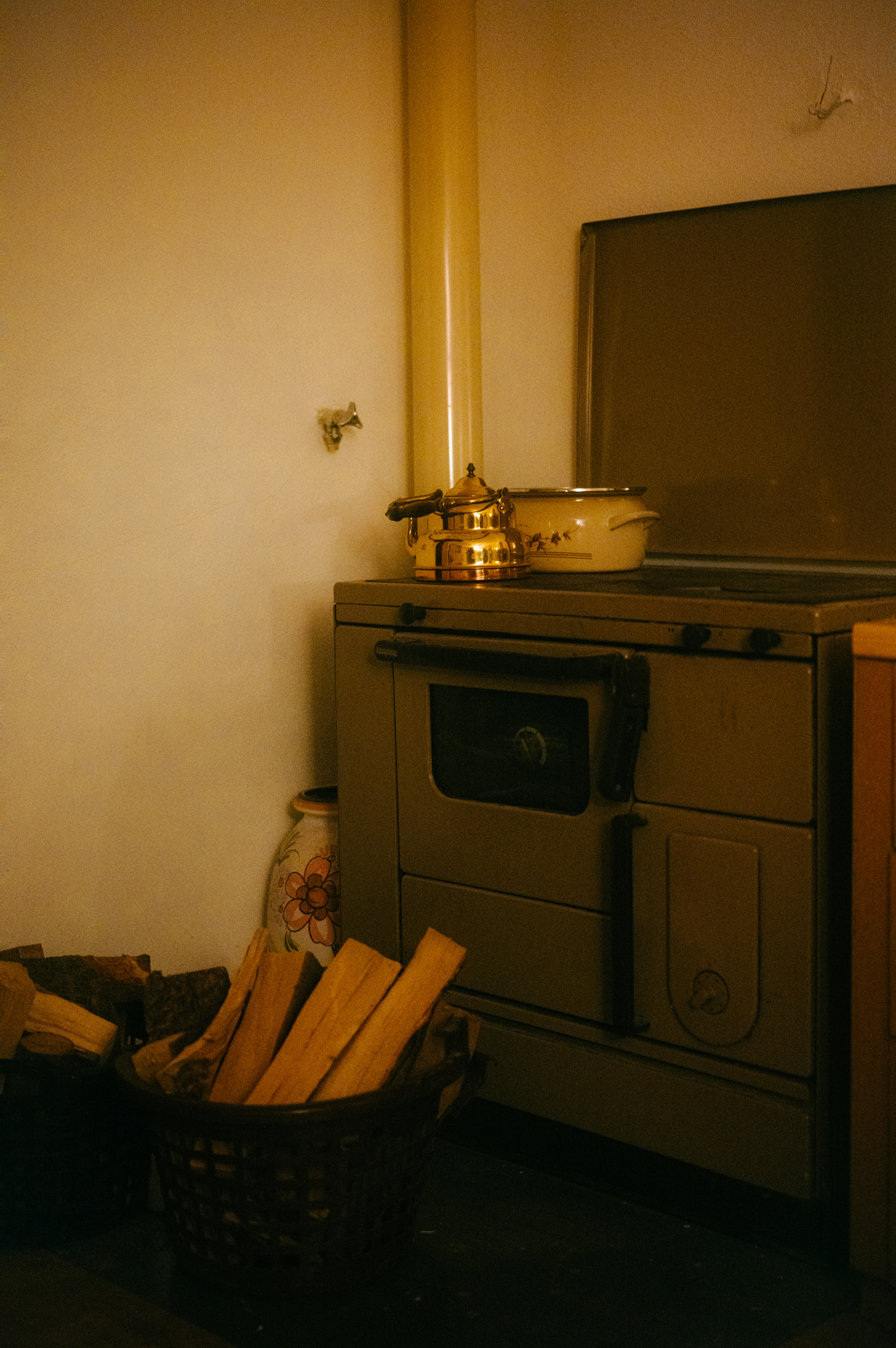 Antique stove with a gleaming kettle and a basket of firewood, evoking a sense of warmth and nostalgia in a rustic kitchen setting.