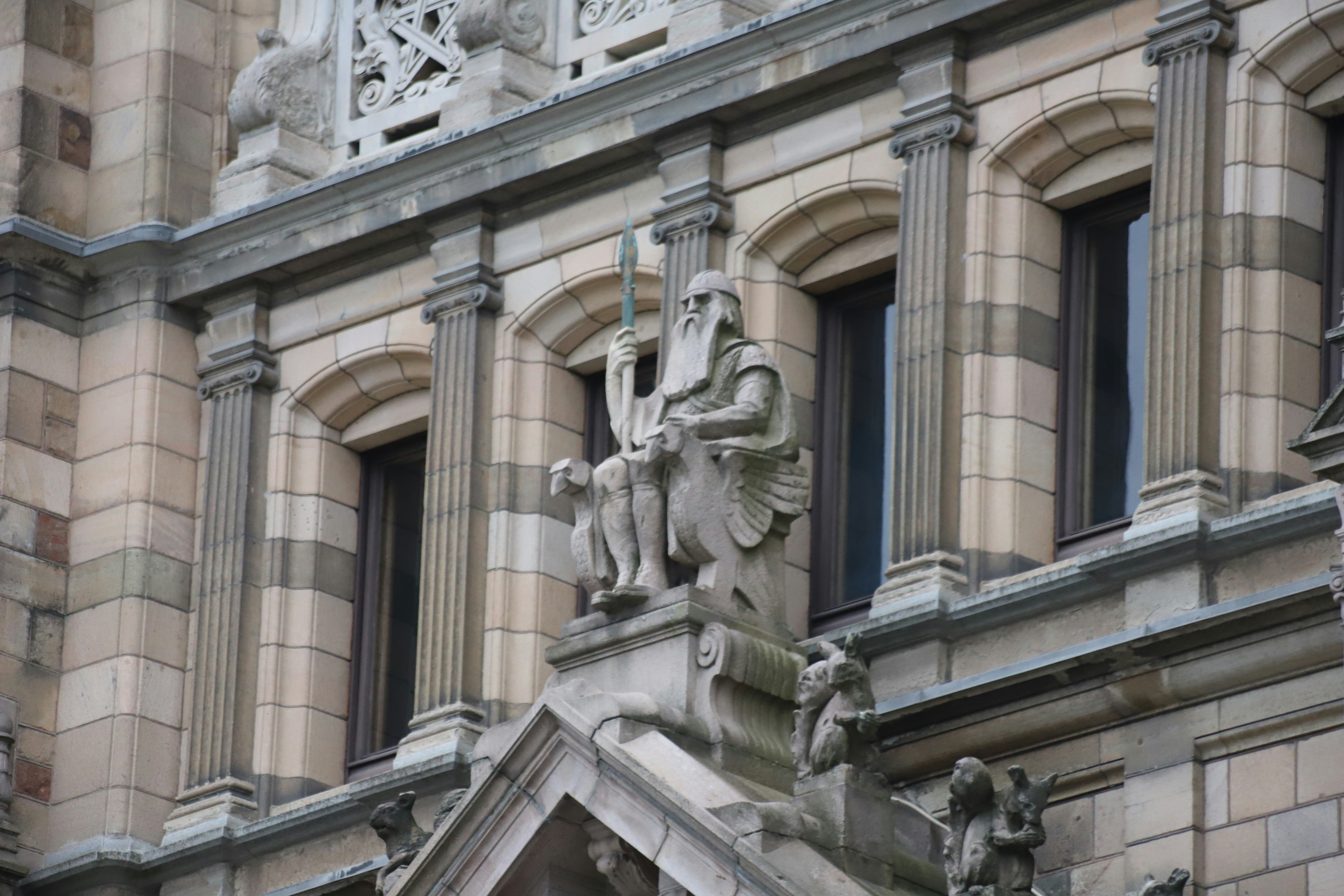 Stone statue of a seated figure holding a staff, set against a classical building facade with arched windows and detailed masonry.