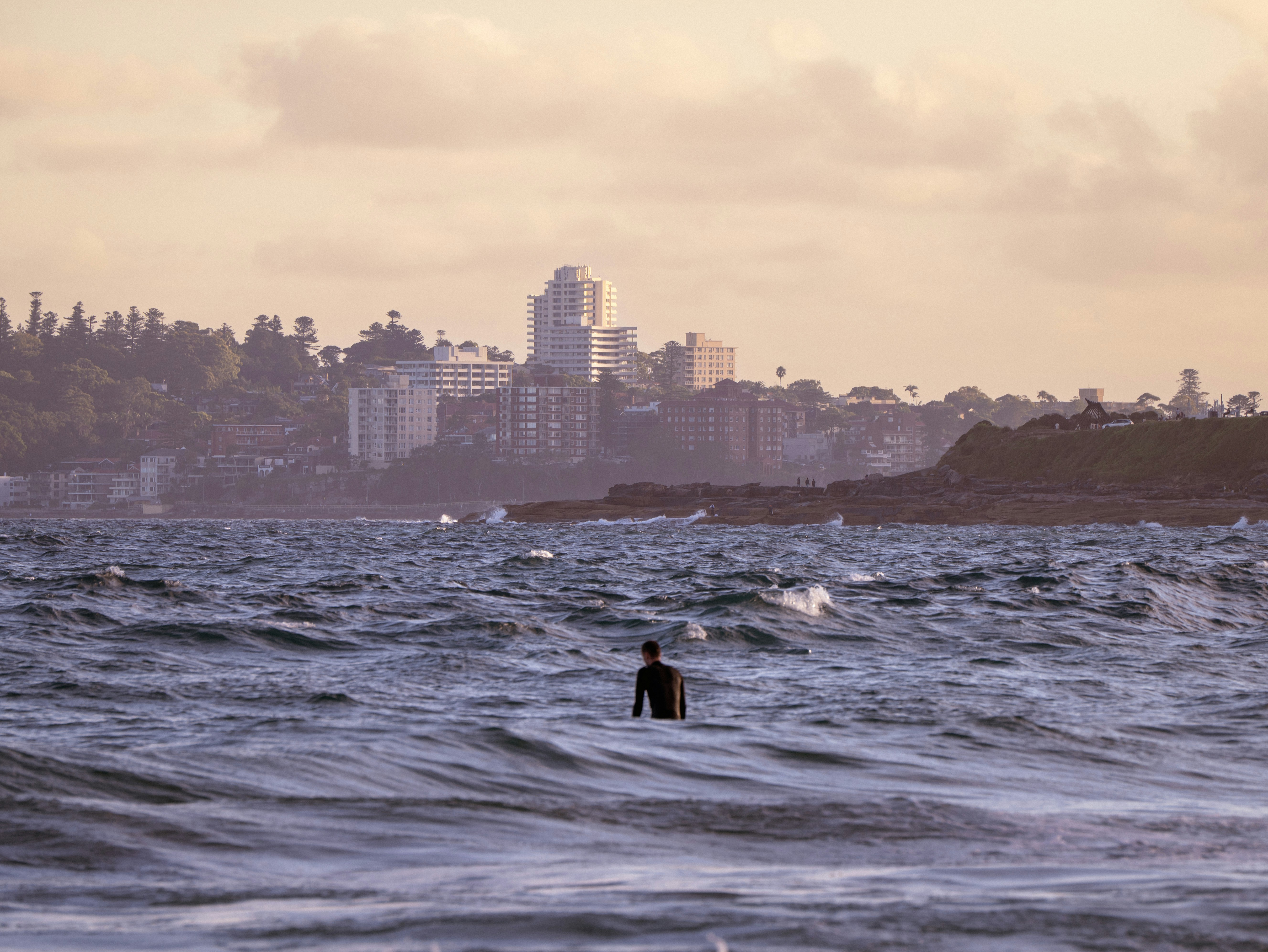 Twilight Silhouette: A Lone Figure in Choppy Seas Beneath a Distant City Skyline