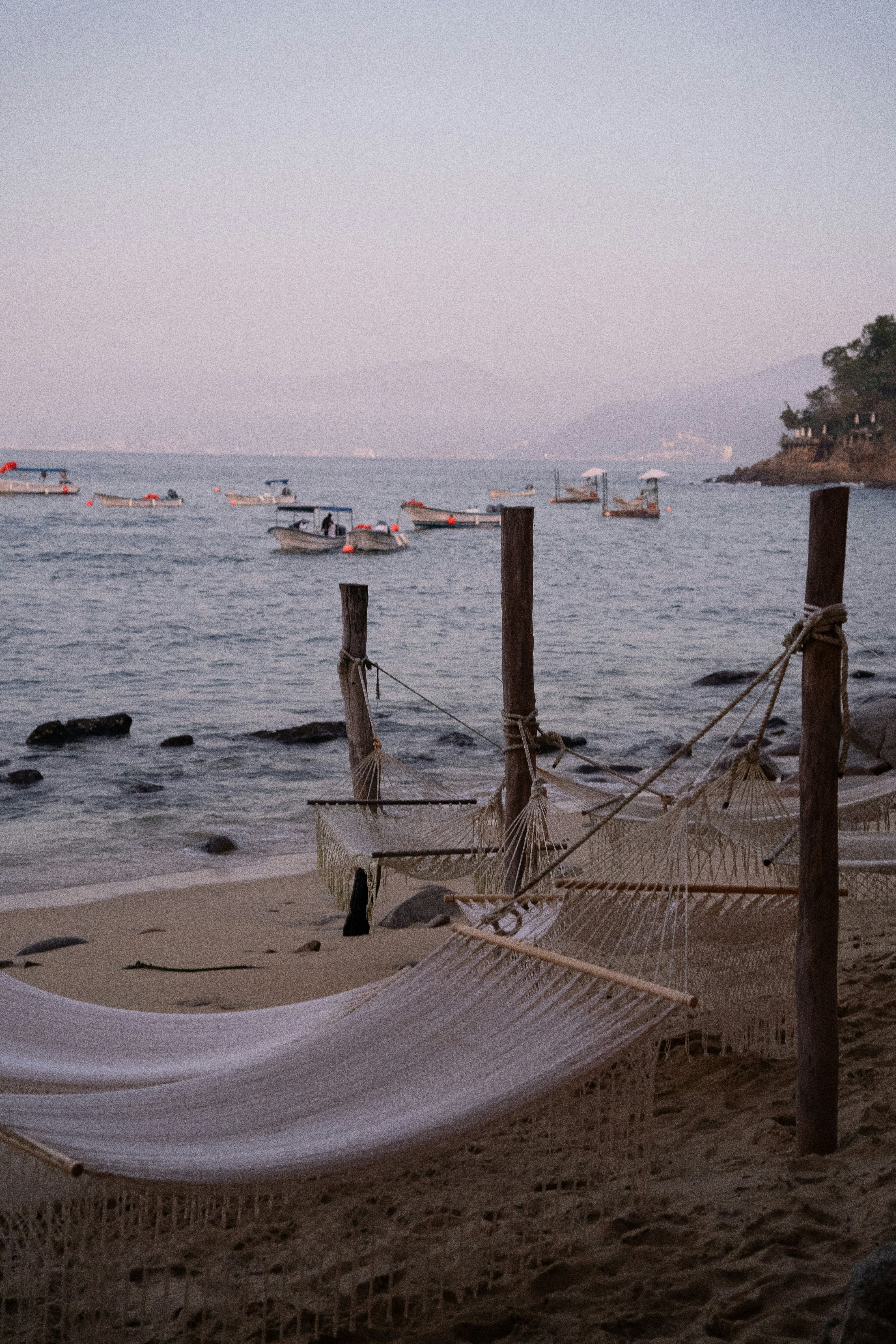 Hammocks hang between wooden posts on a sandy beach with boats dotting the calm ocean in the background.