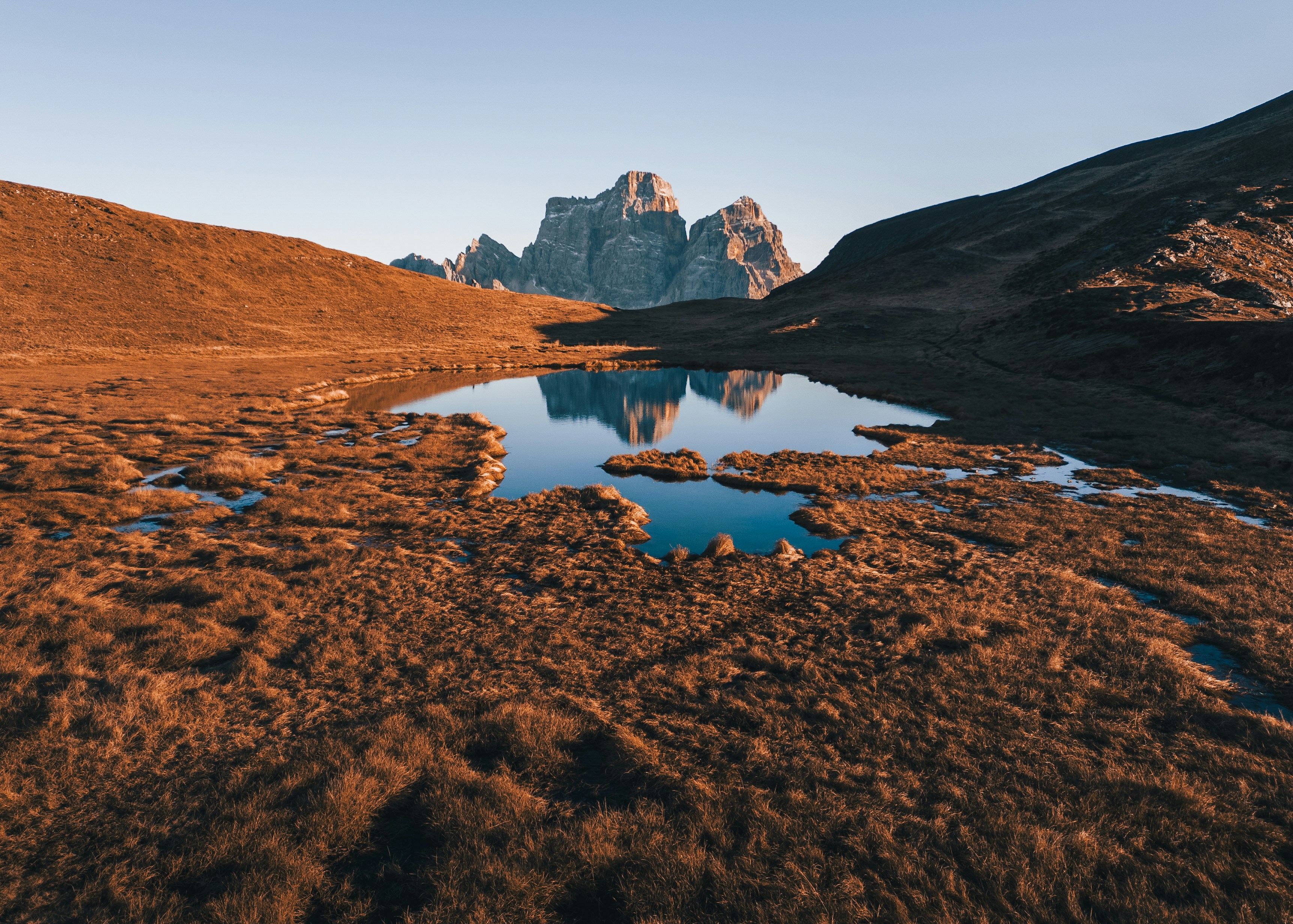 Mountain peaks reflected in a serene lake surrounded by warm golden terrain under a clear blue sky.