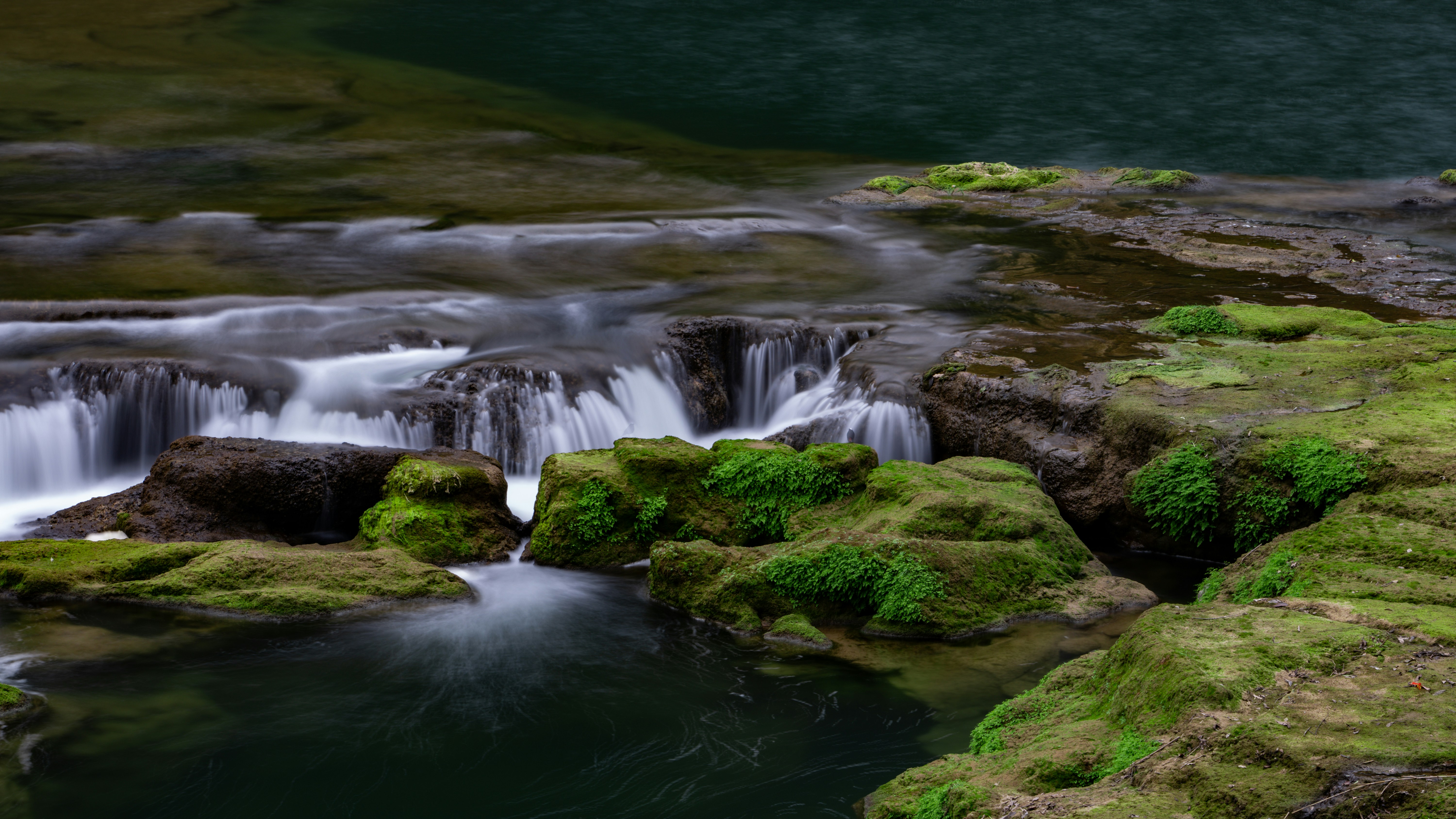 Small waterfall cascading over moss-covered rocks into a calm pool surrounded by greenery.