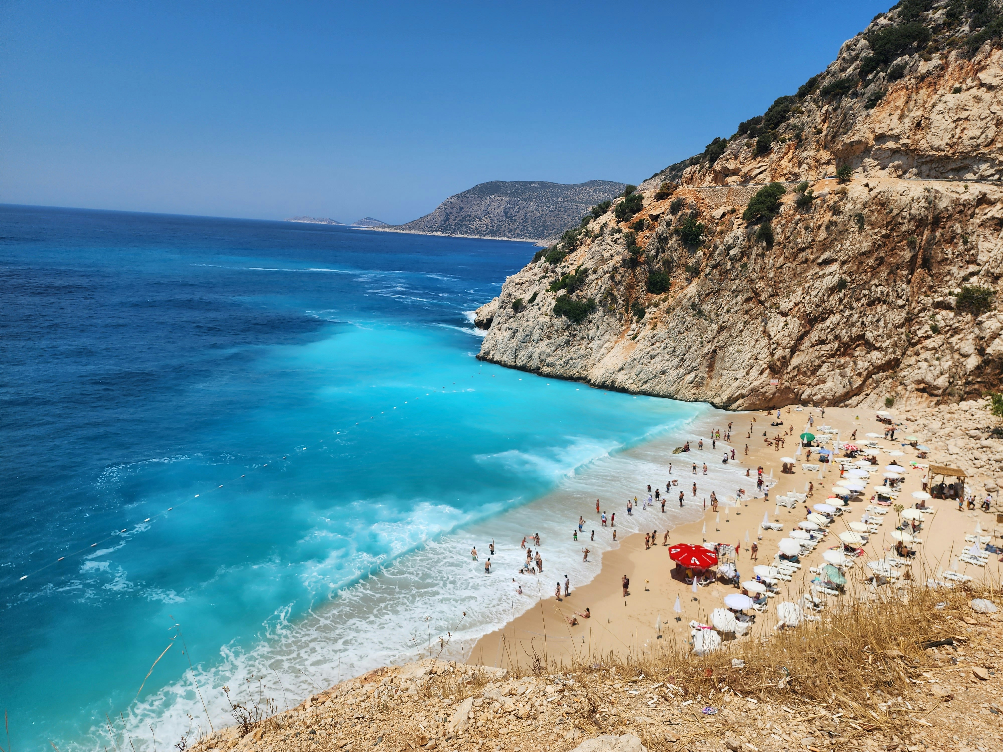 Secluded beach with turquoise waters and sunbathers under umbrellas, nestled between rocky cliffs.