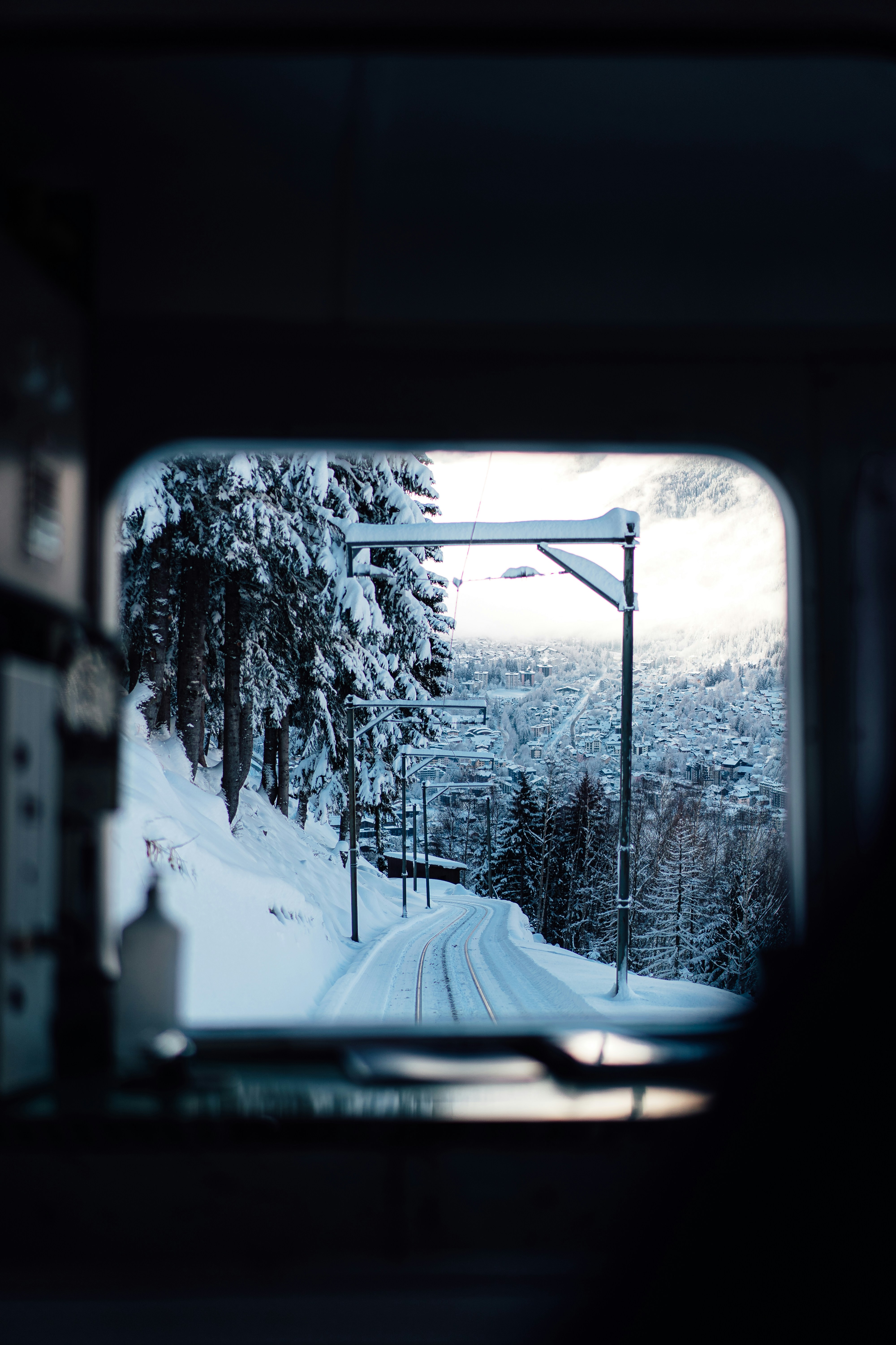 Snow-covered railway framed by a train window, leading through a frosted forest into a distant town under soft morning light.