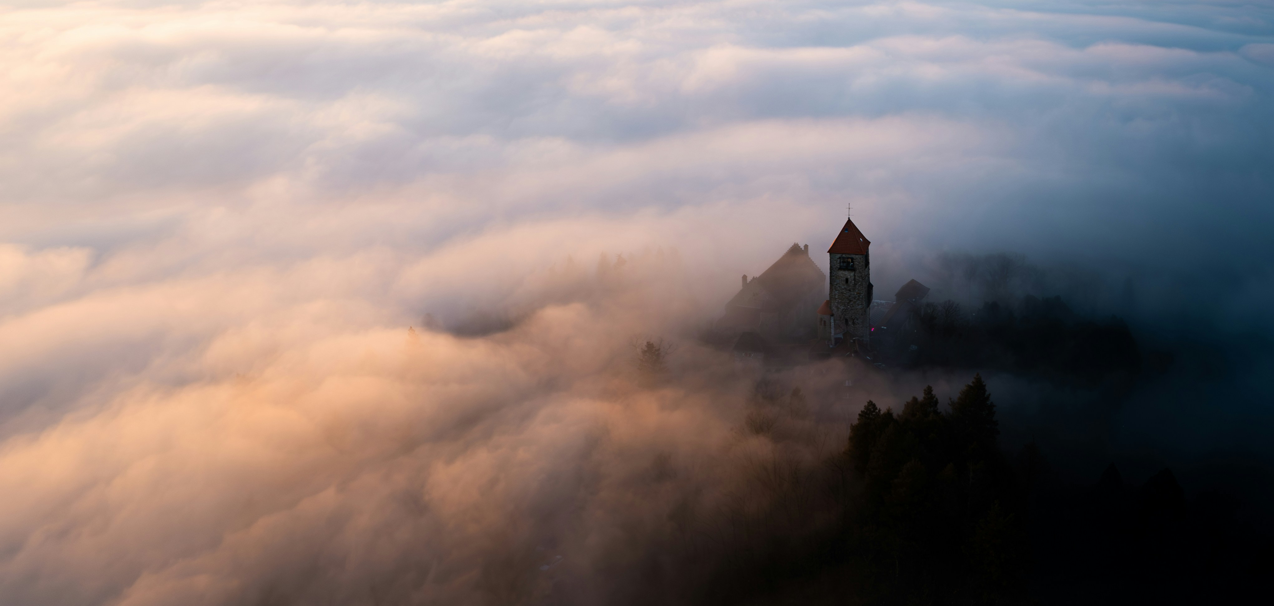 An aerial view of a stone tower with an orange roof surrounded by dense fog and mist during sunrise. The light casts a warm glow on the clouds, creating a soft and serene atmosphere. Trees and additional structures are partially visible through the mist.