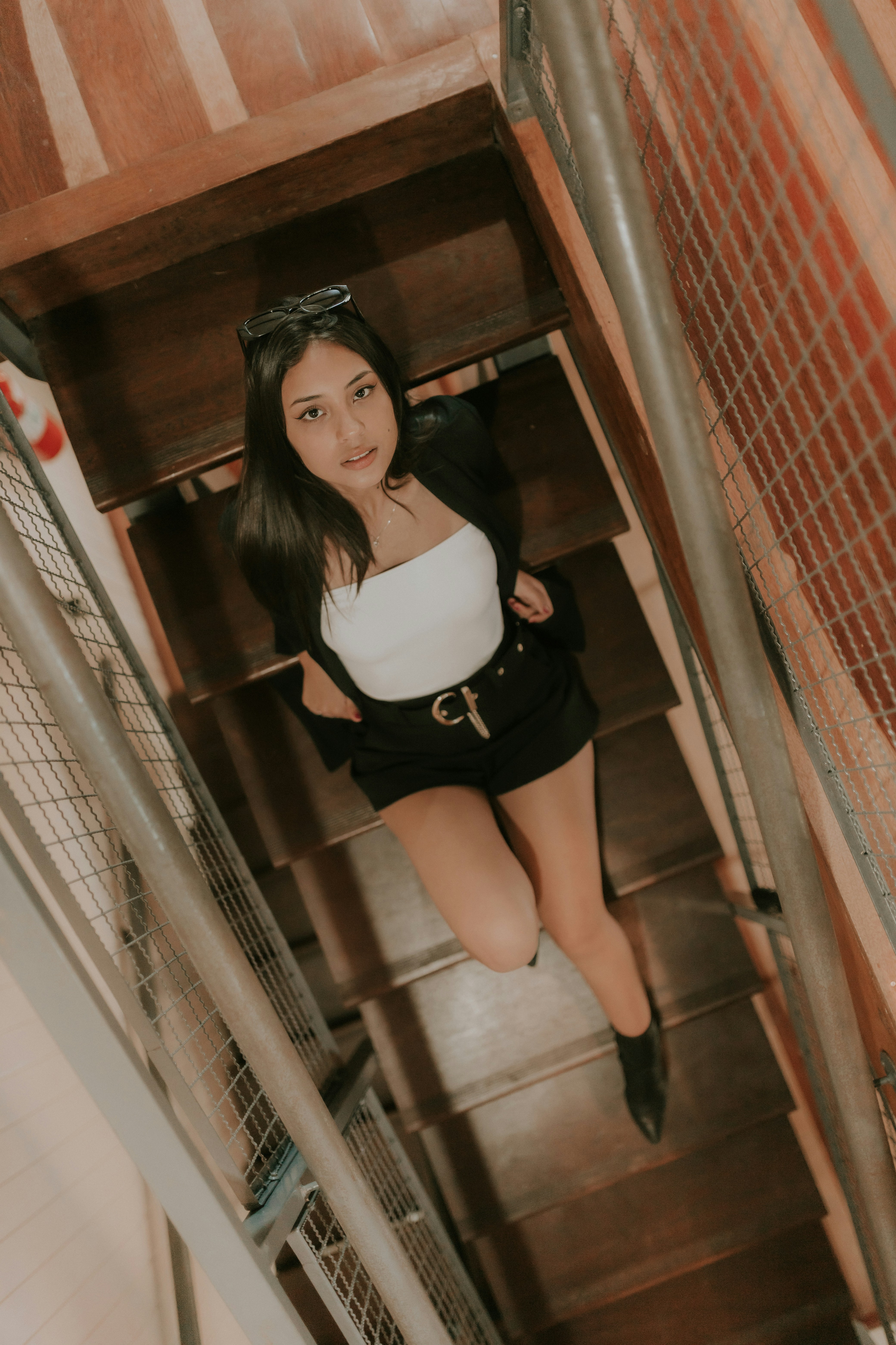 A young woman in a chic outfit poses on wooden stairs, exuding confidence and style. Her gaze draws the viewer in, highlighting the modern interior setting.