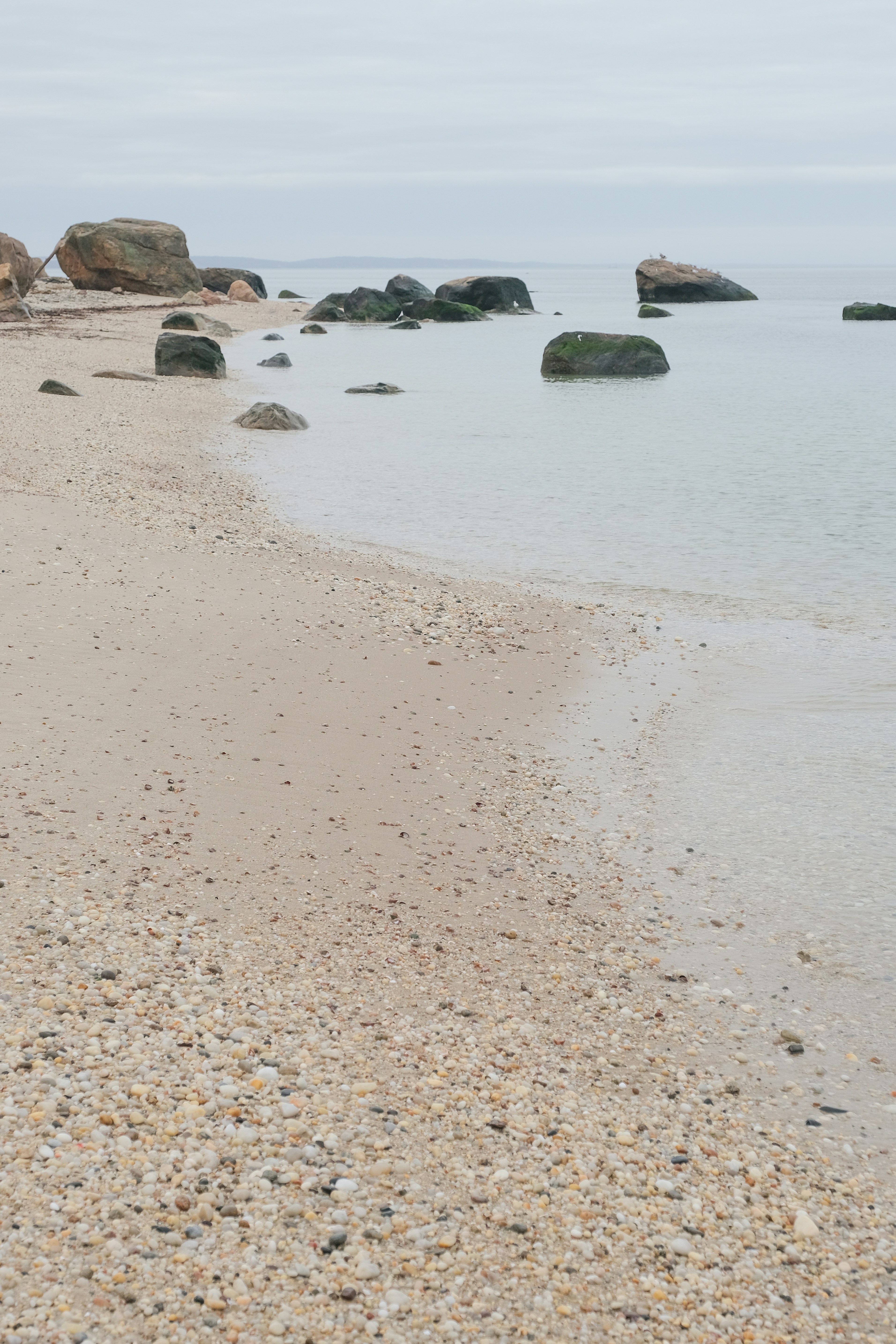 A quiet beach landscape with large boulders and a calm ocean under an overcast sky.