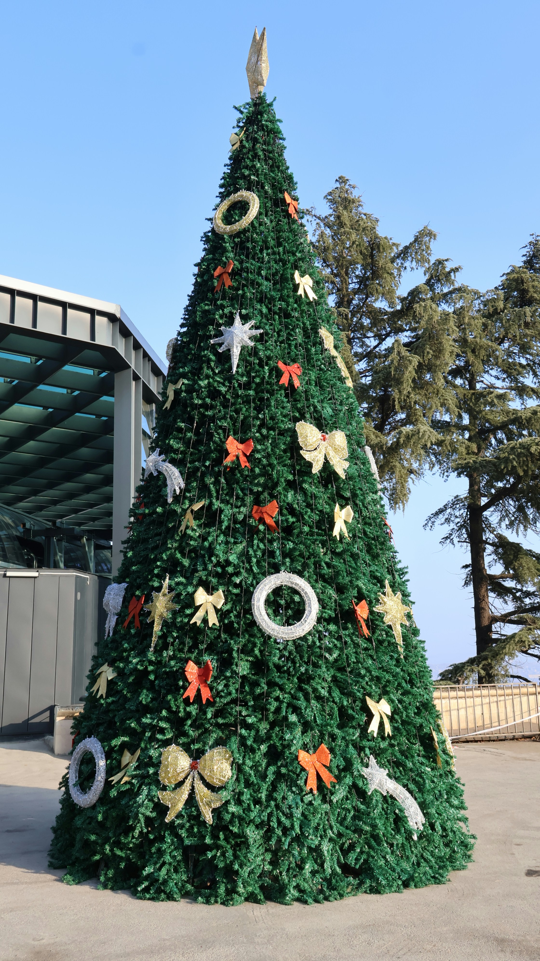 Vertical Holiday Spire: A Verdant Christmas Tree Adorned with Ribbons, Wreaths, and Ornaments by a Modern Plaza