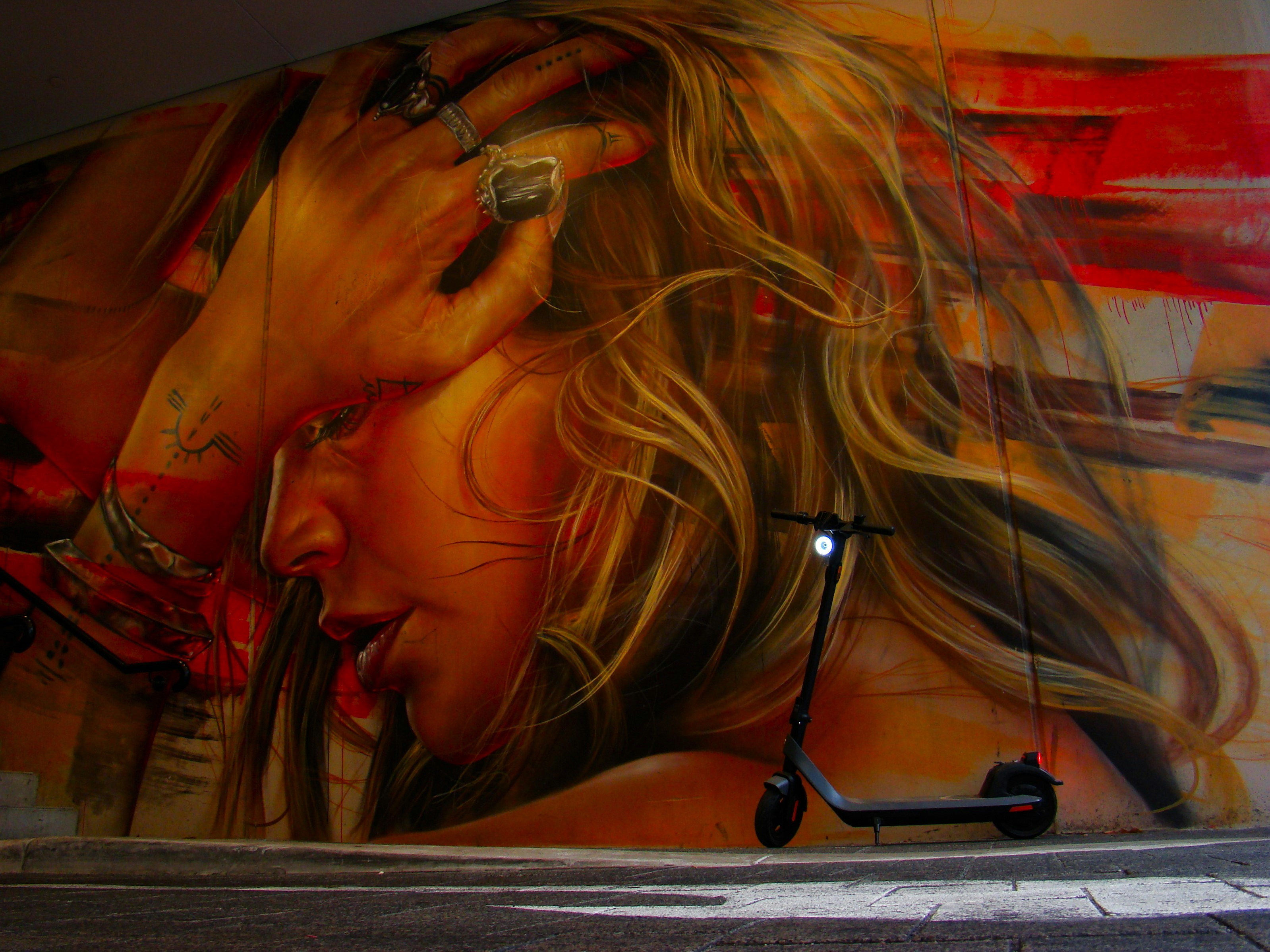 Wall mural of a woman with wind-swept hair in warm reds and golds, set against an urban backdrop. A small electric scooter sits in front, partially obscuring the artwork.