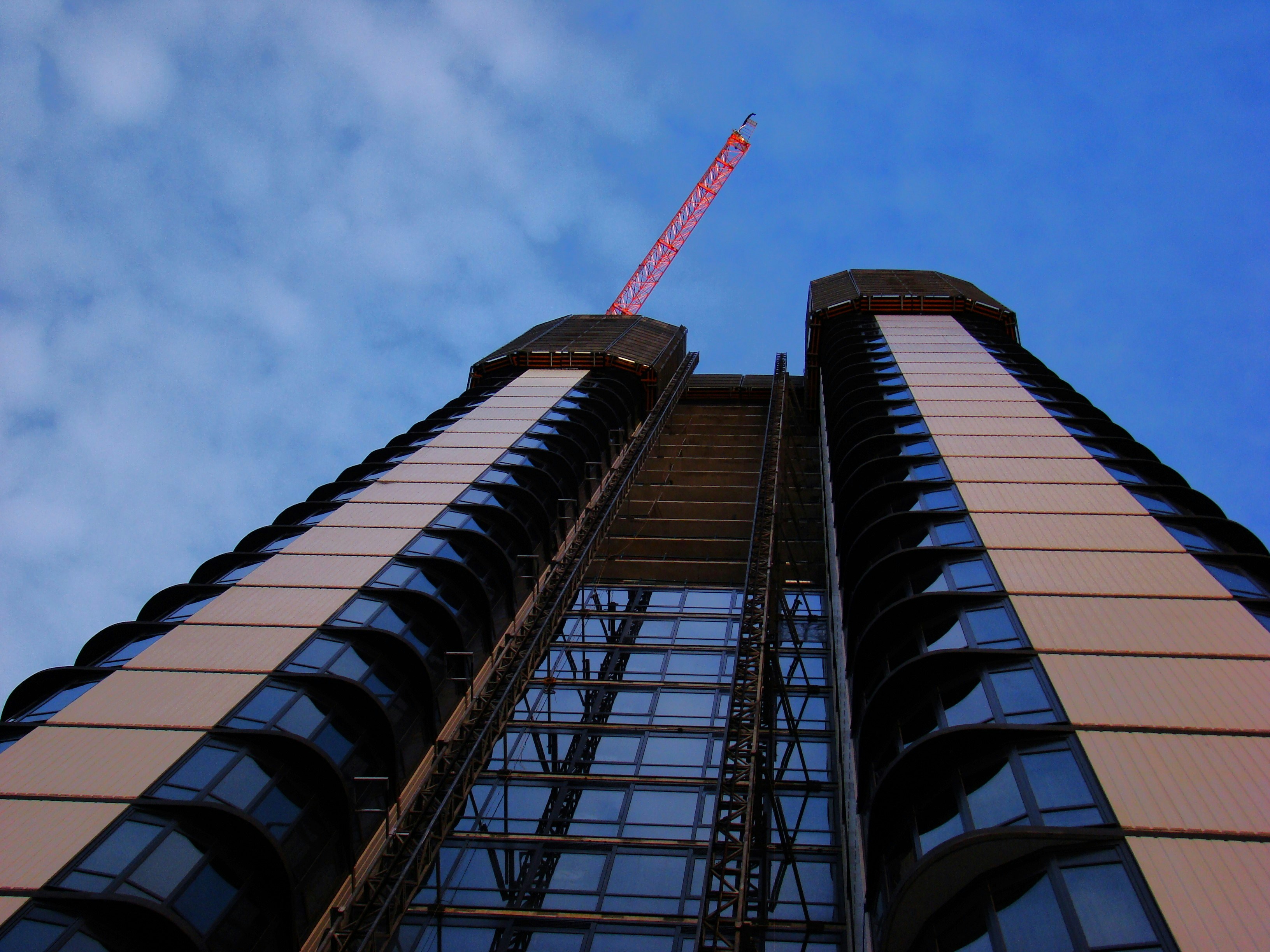 Low-angle shot of two glass-clad towers under construction, with a red crane stretching between them against a clear blue sky.