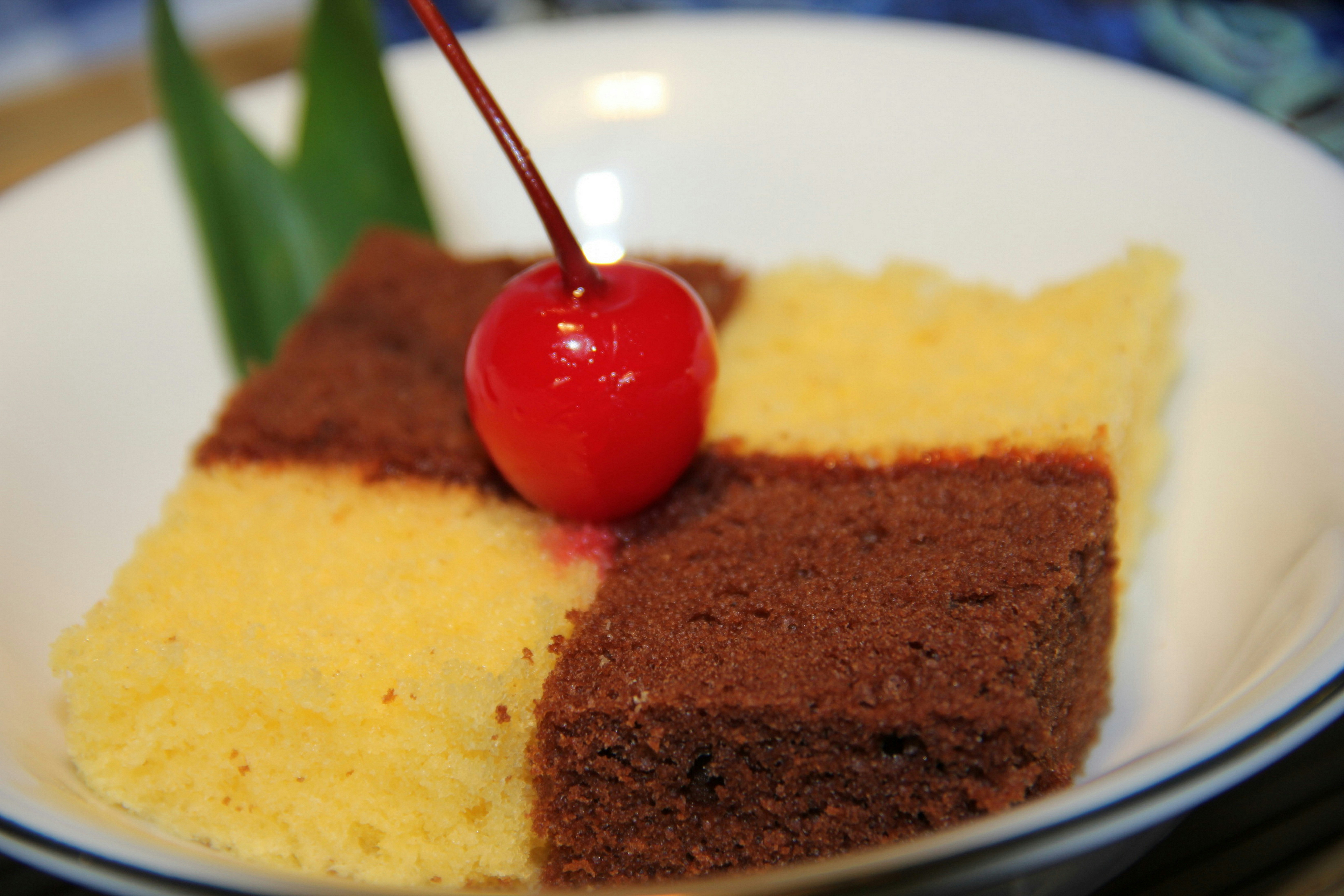 Four distinct cake squares, alternating between yellow and brown, topped with a bright red cherry, set against a simple backdrop.
