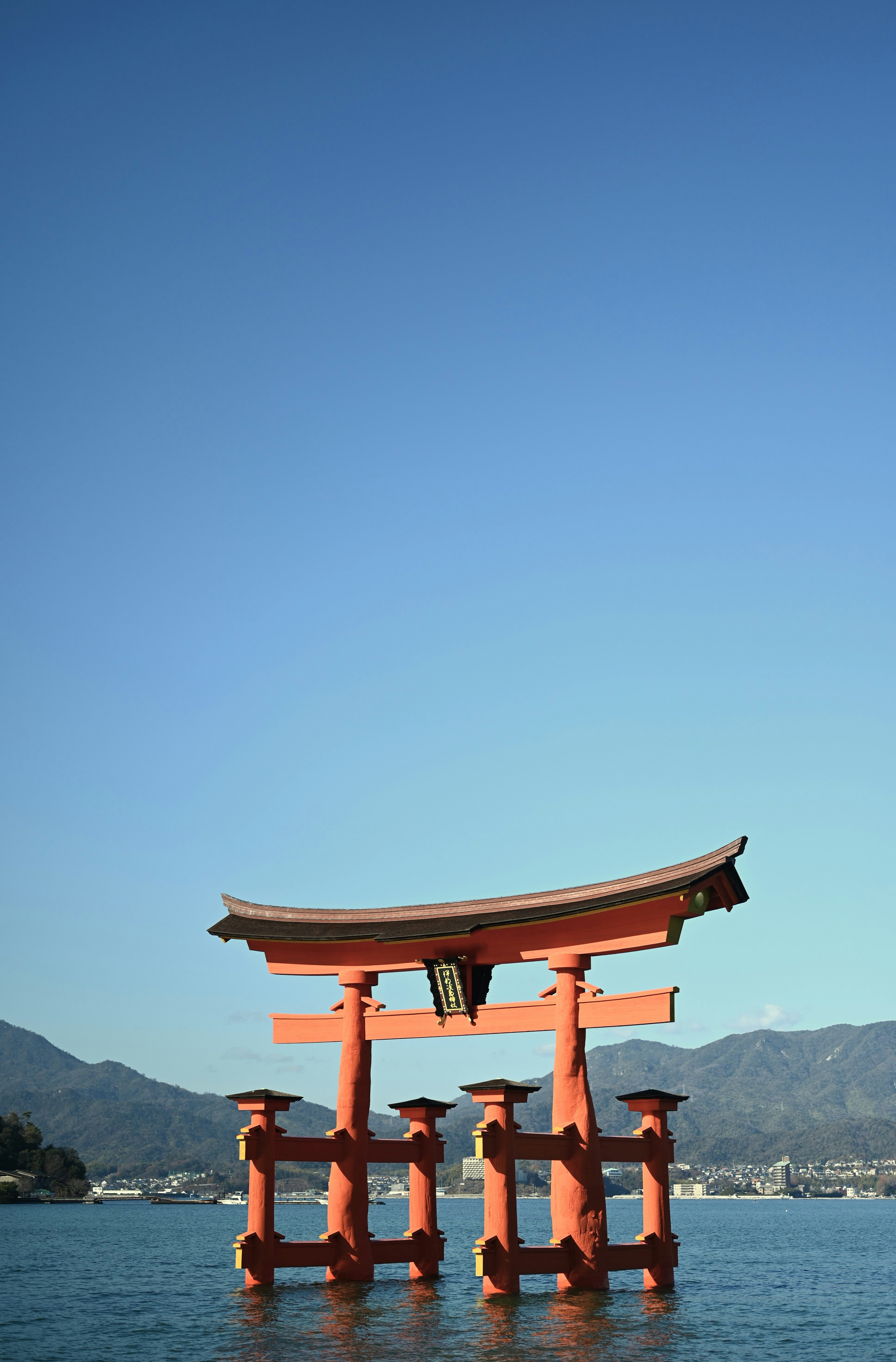 A vibrant red torii gate stands majestically in calm waters, framed by distant mountains and a clear blue sky.