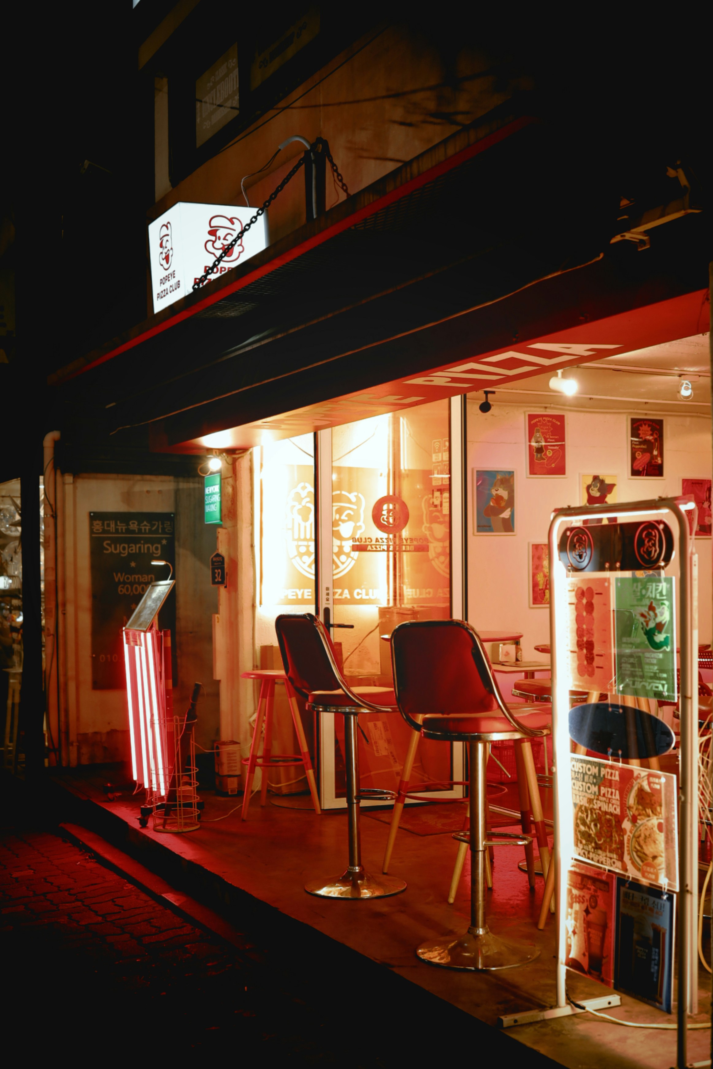 Retro-themed pizza restaurant with red and orange neon lights and high bar stools outside.