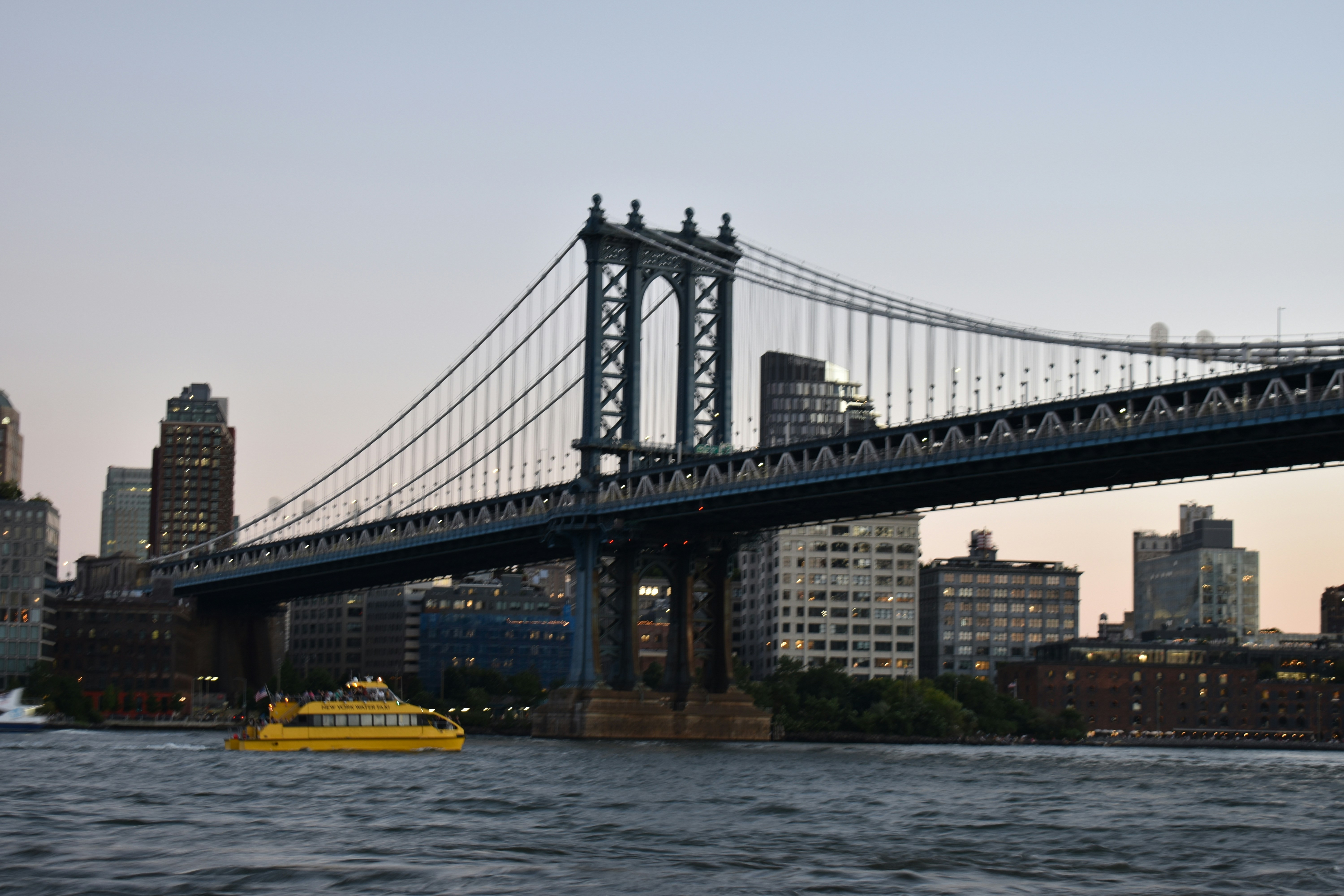 A yellow water taxi is driving under the Manhattan Bridge, New York, at sunset.