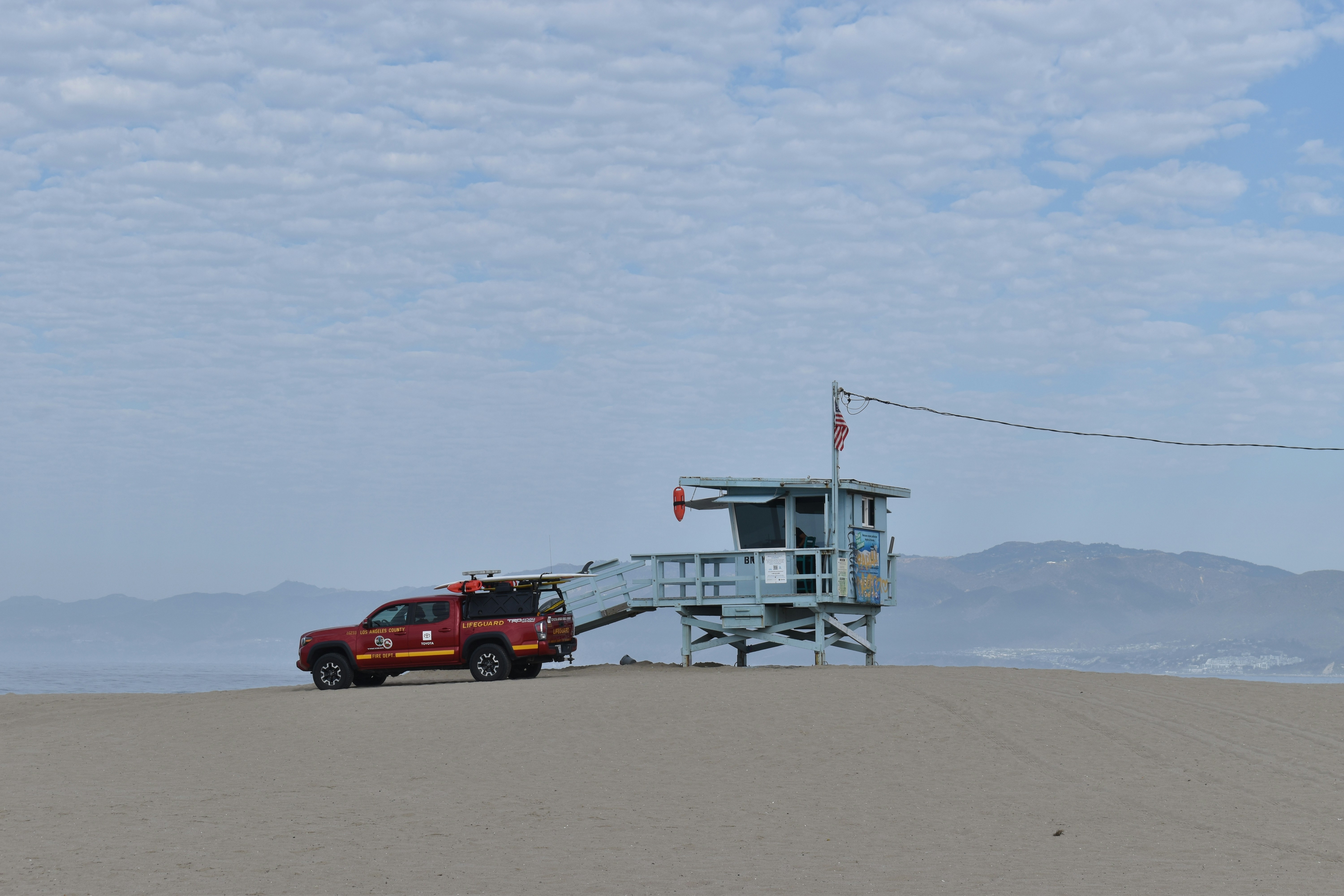 A lifeguard station with a red pick up truck parked on the sand in front of it with the Santa Monica mountains in the background.