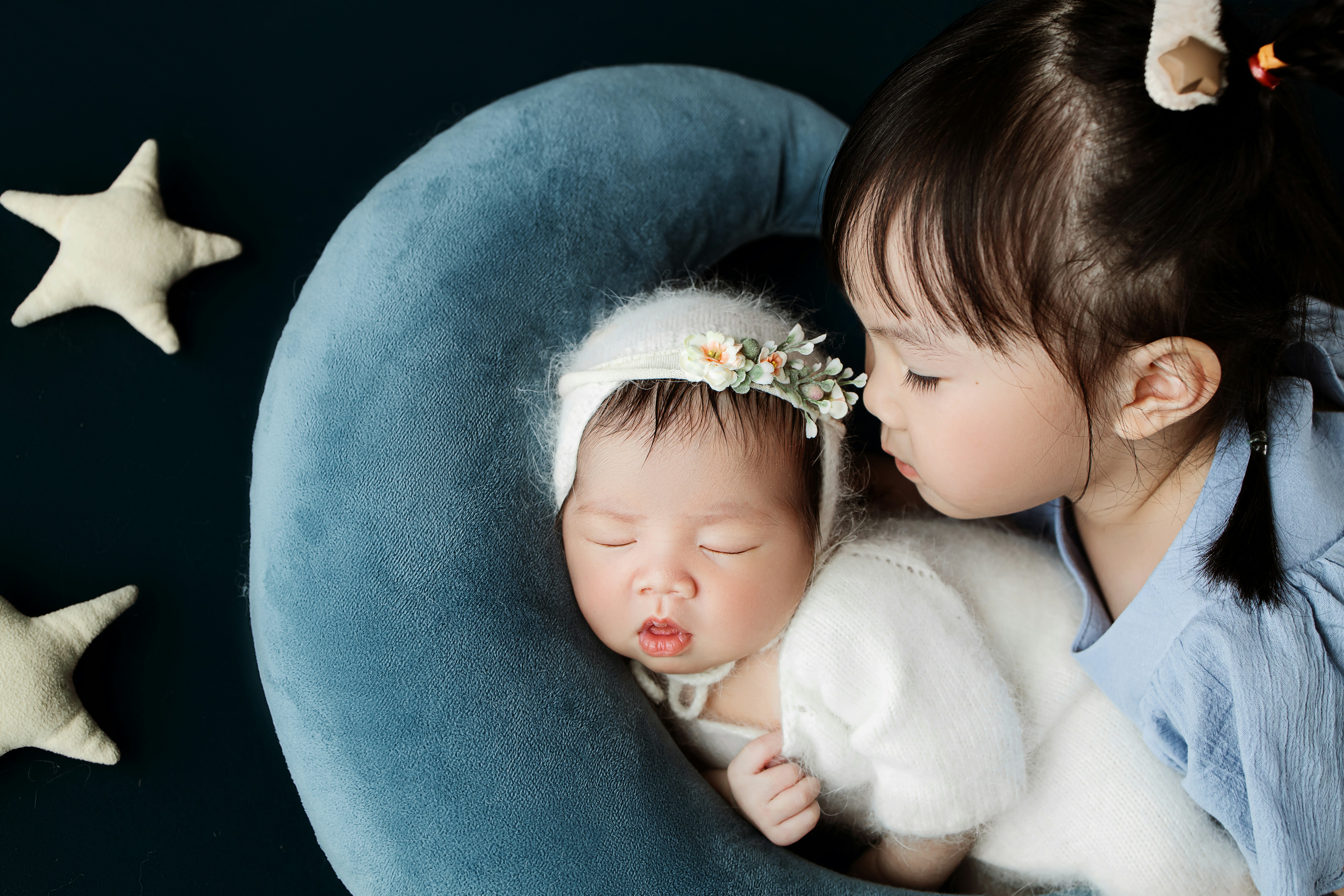 Young child gently kisses a sleeping baby nestled in a crescent moon cushion surrounded by stars.