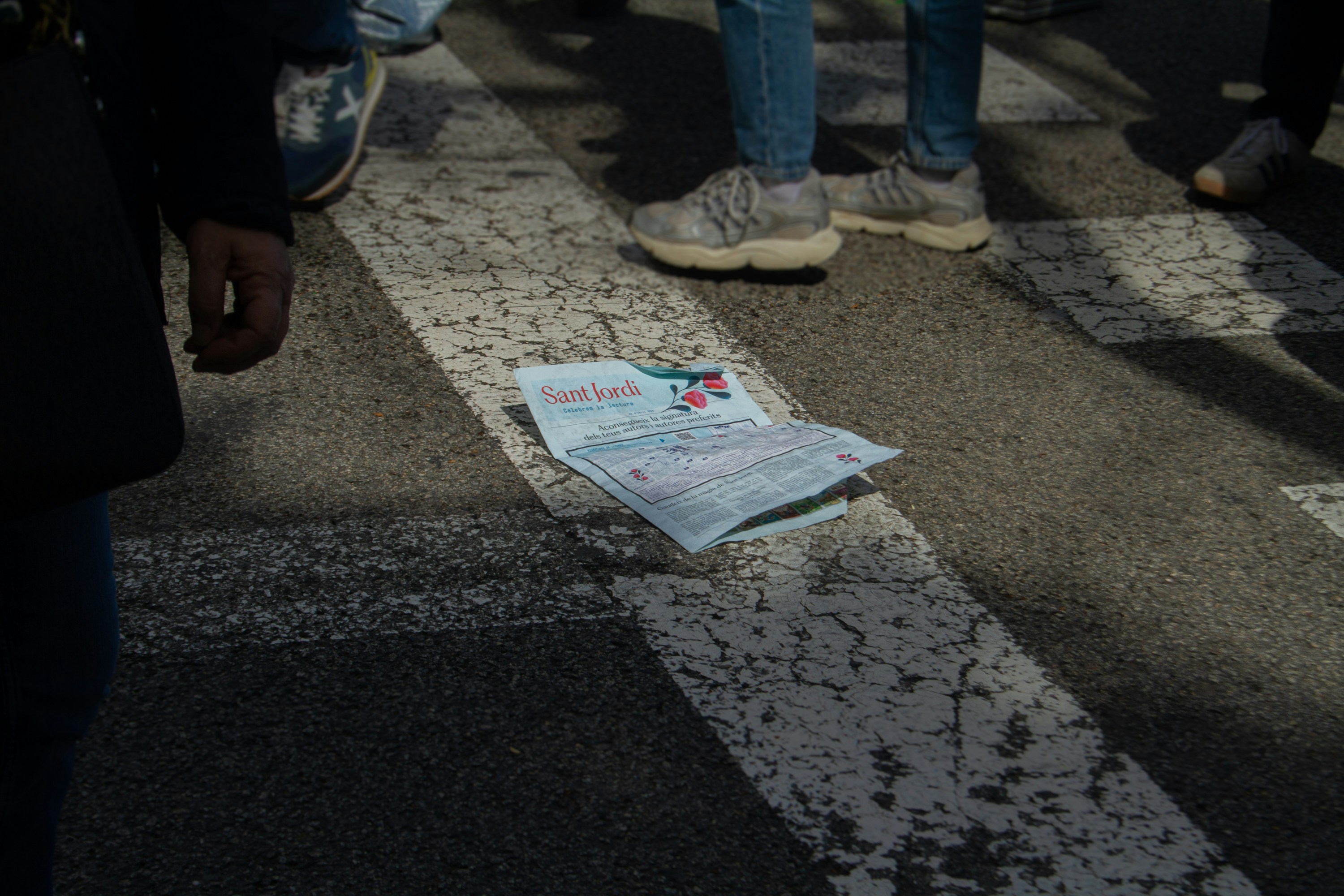 Discarded paper on a crosswalk surrounded by pedestrians' feet in sunlight.