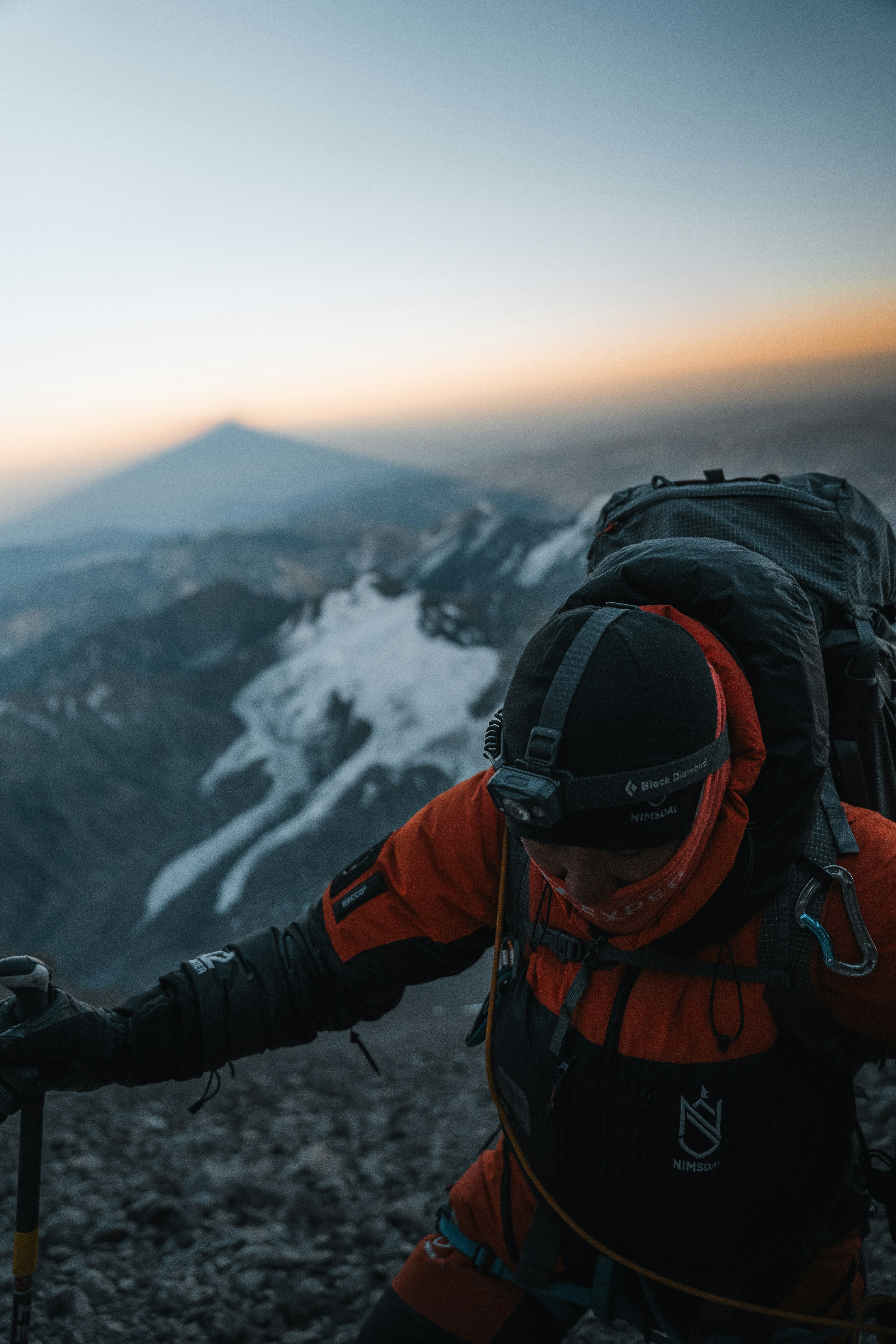 Mountaineer in red gear climbs rocky terrain with snow-covered peaks and a dawn-lit sky in the background.