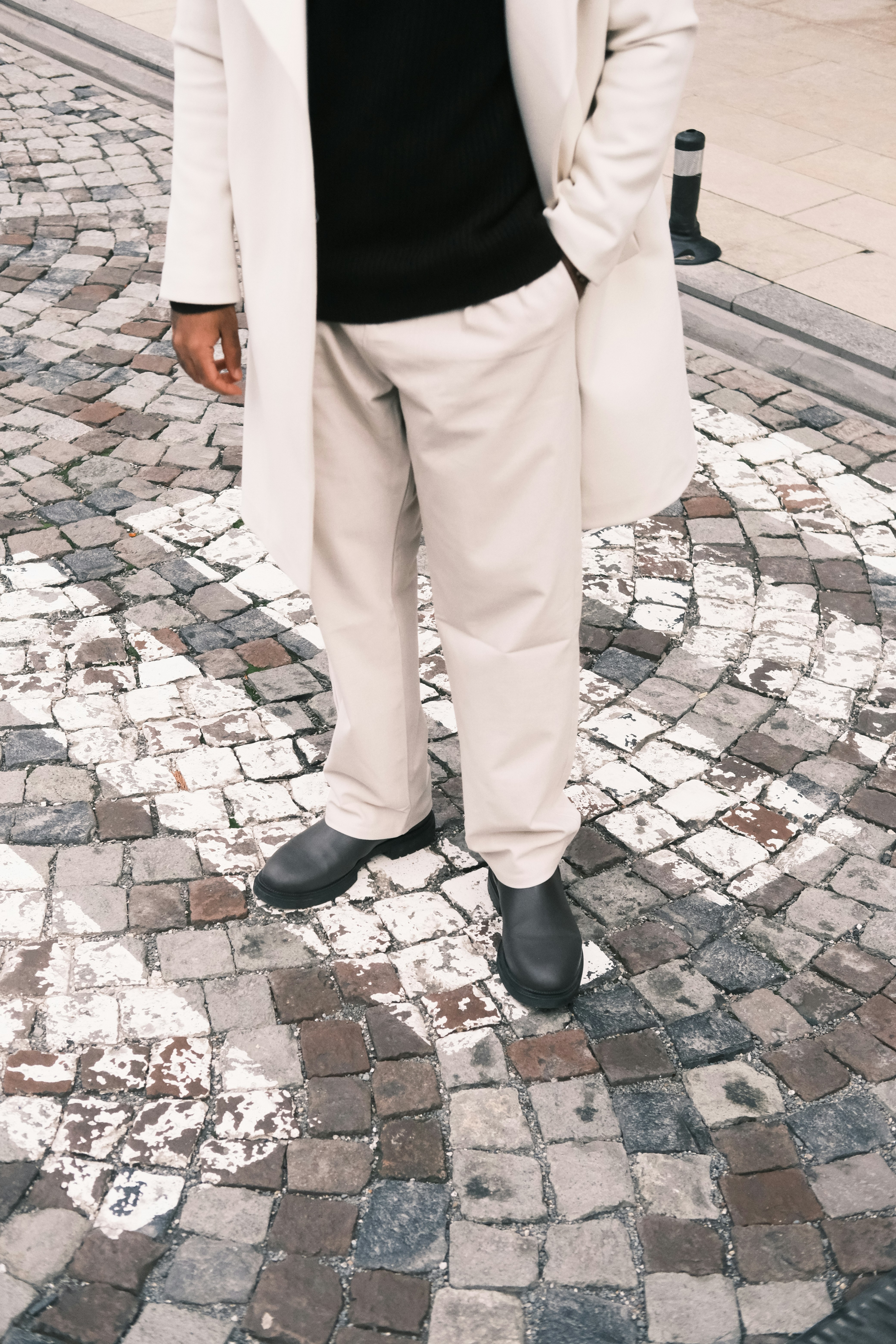 A fashionable individual dressed in a cream coat and light trousers stands confidently on a cobblestone path featuring a circular design.
