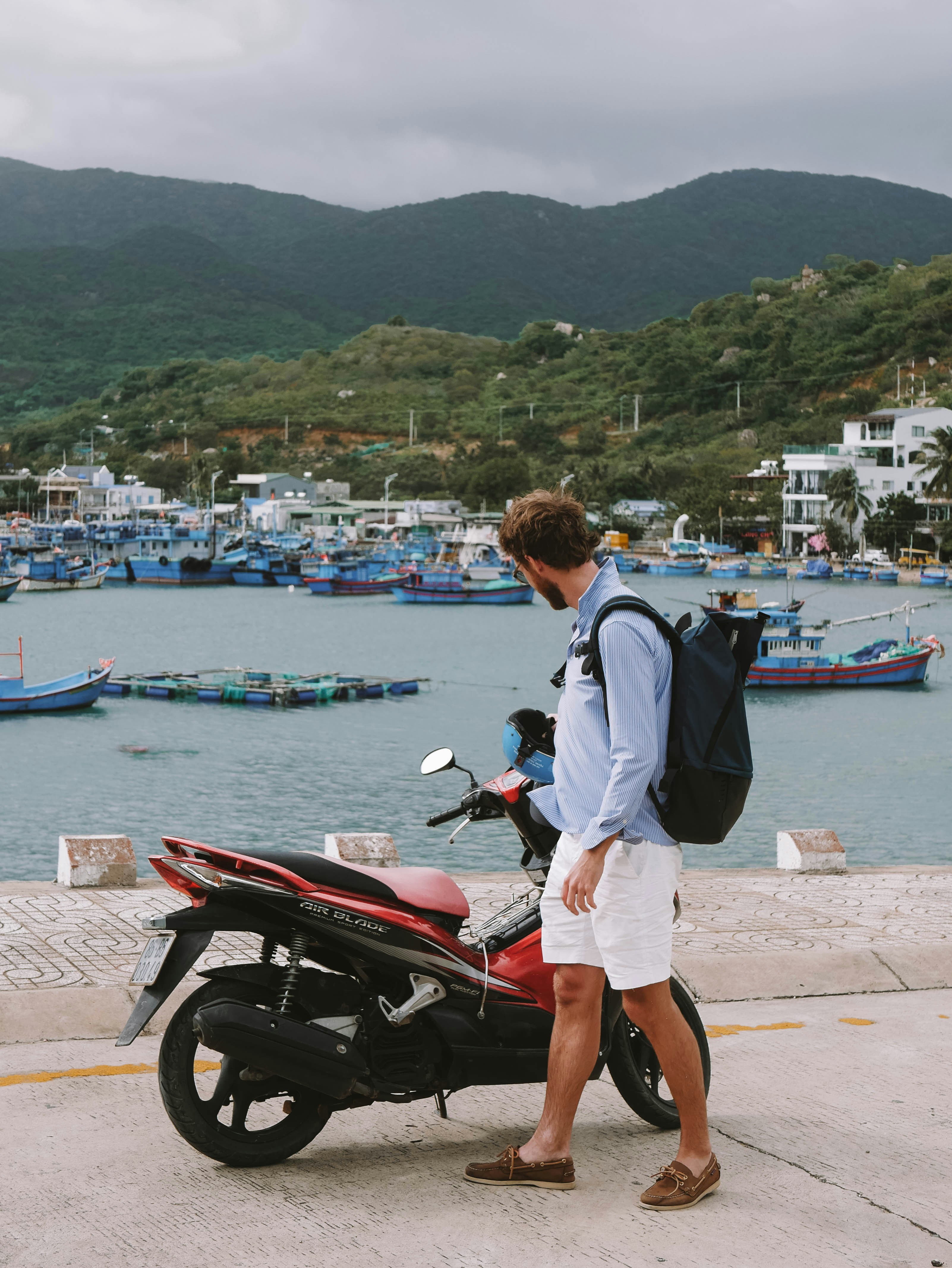 Harbor Pause: A Backpacked Wanderer Pauses by a Red Scooter Over a Quiet Marina