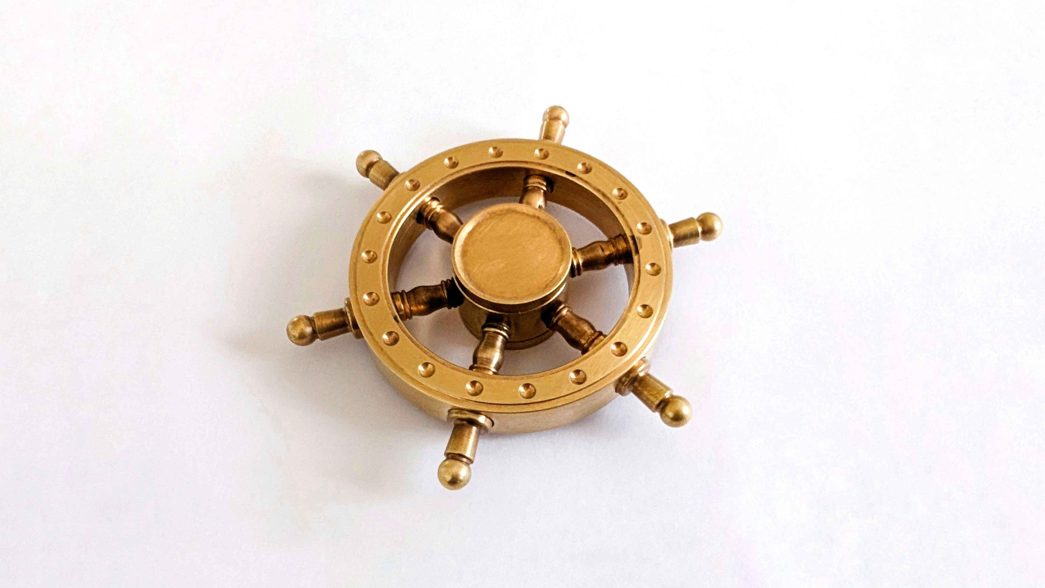 Close-up of a brass ship wheel resting on a clean white background. The photograph highlights the wheel's riveted spokes and warm metallic texture.