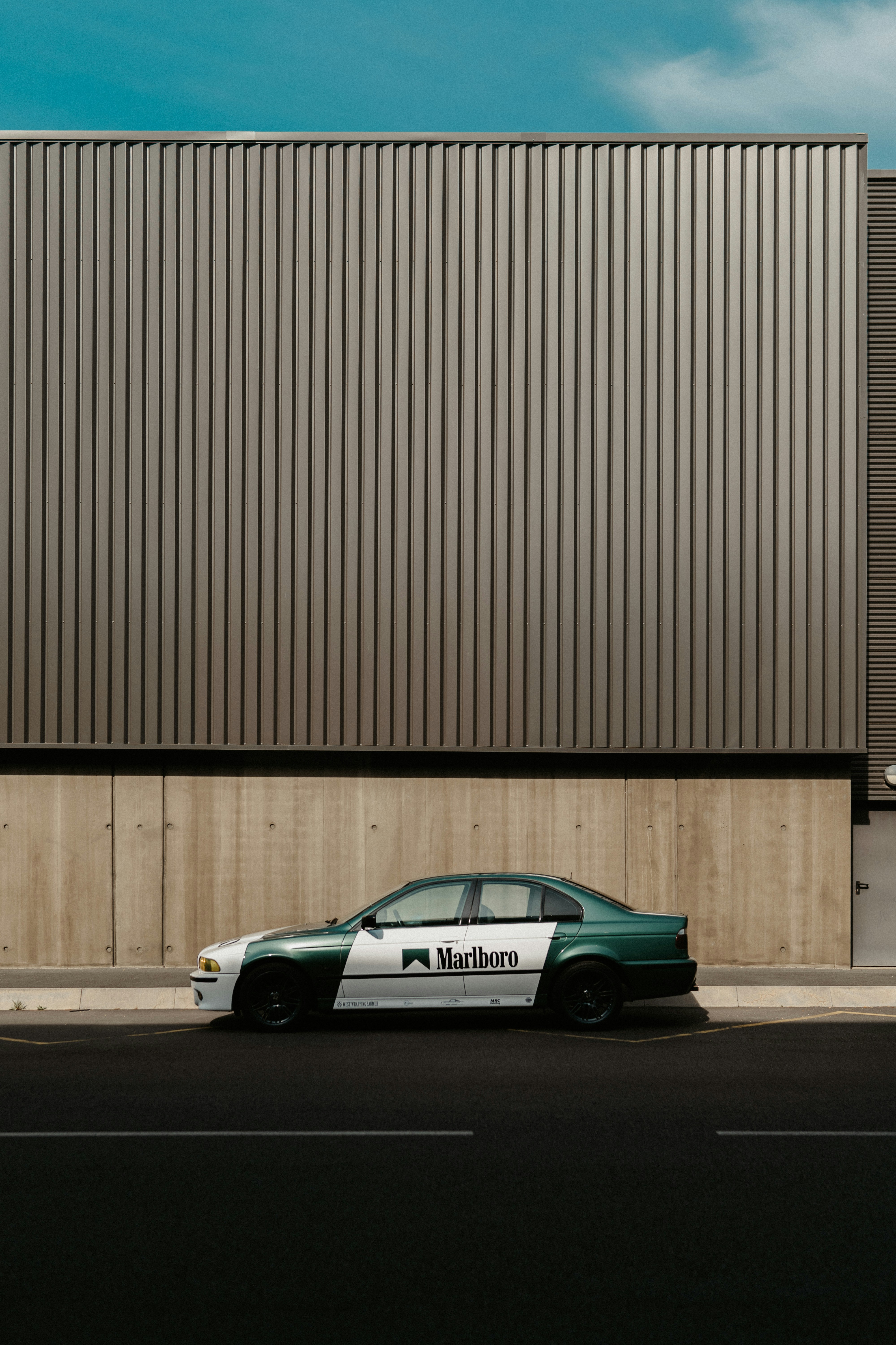 Green and white retro car parked against a modern industrial building with a vertical metal facade under a clear blue sky.