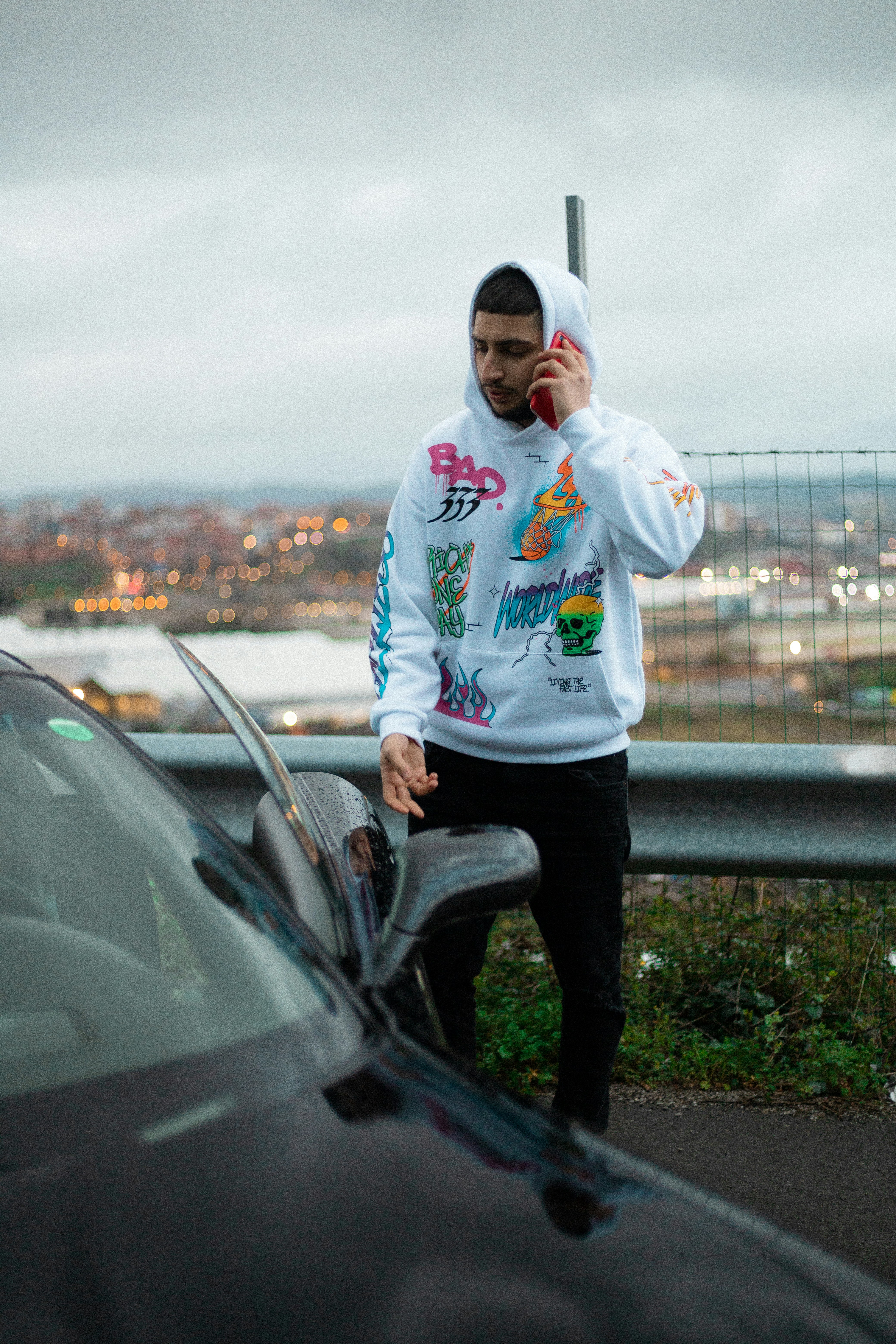 Man in a colorful hoodie talks on the phone next to a car on a city overlook.