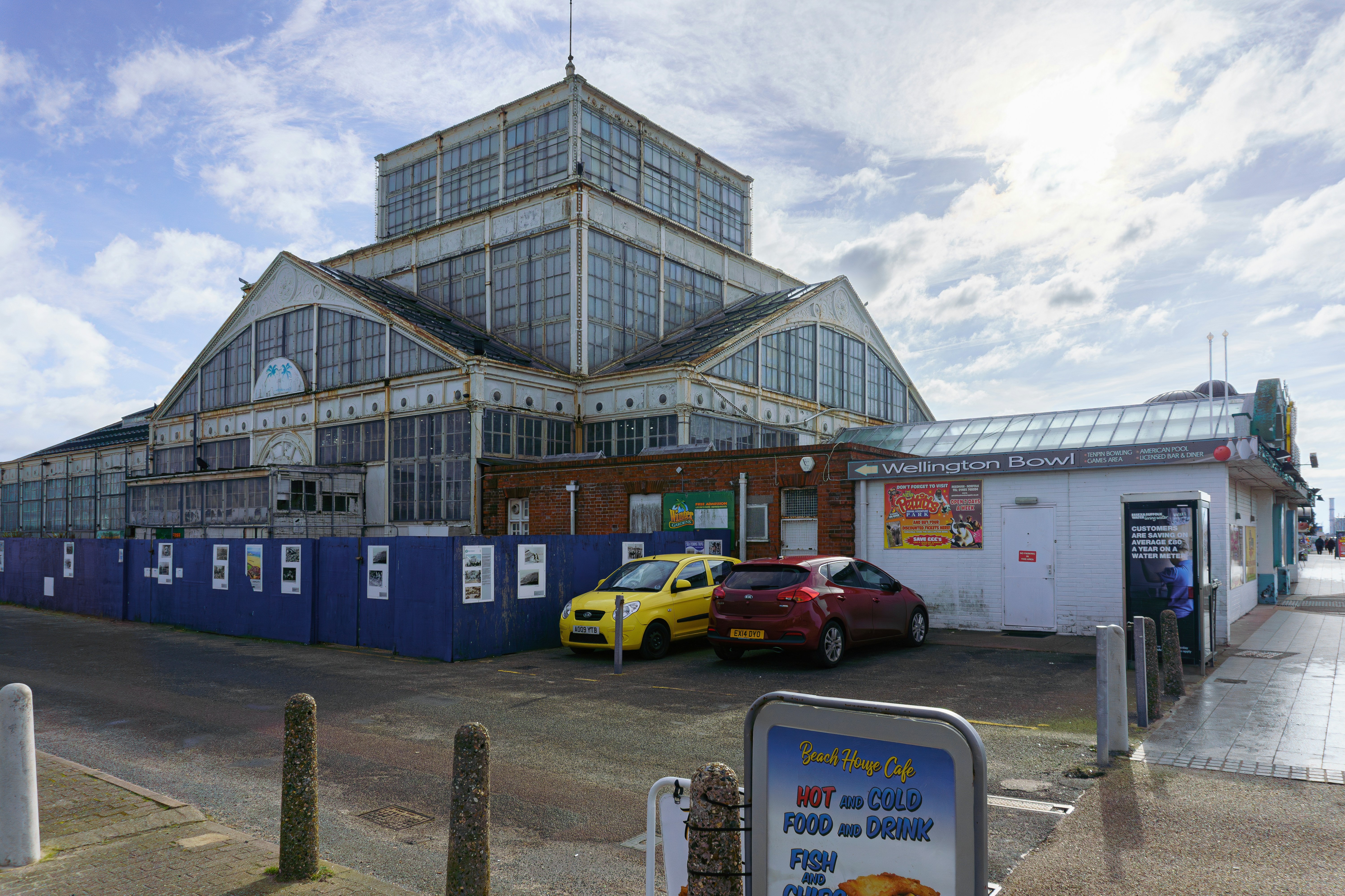 Historic glass-roofed building with surrounding blue construction barriers under a partly cloudy sky.