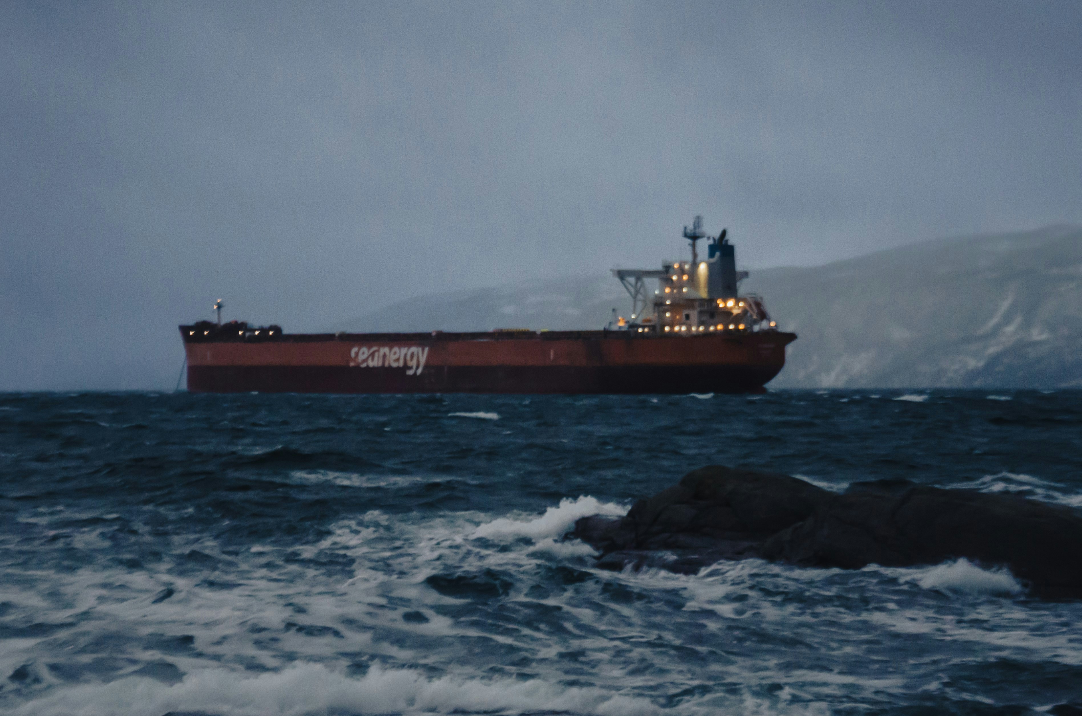 Cargo ship navigating choppy waters under a cloudy sky with distant hills.