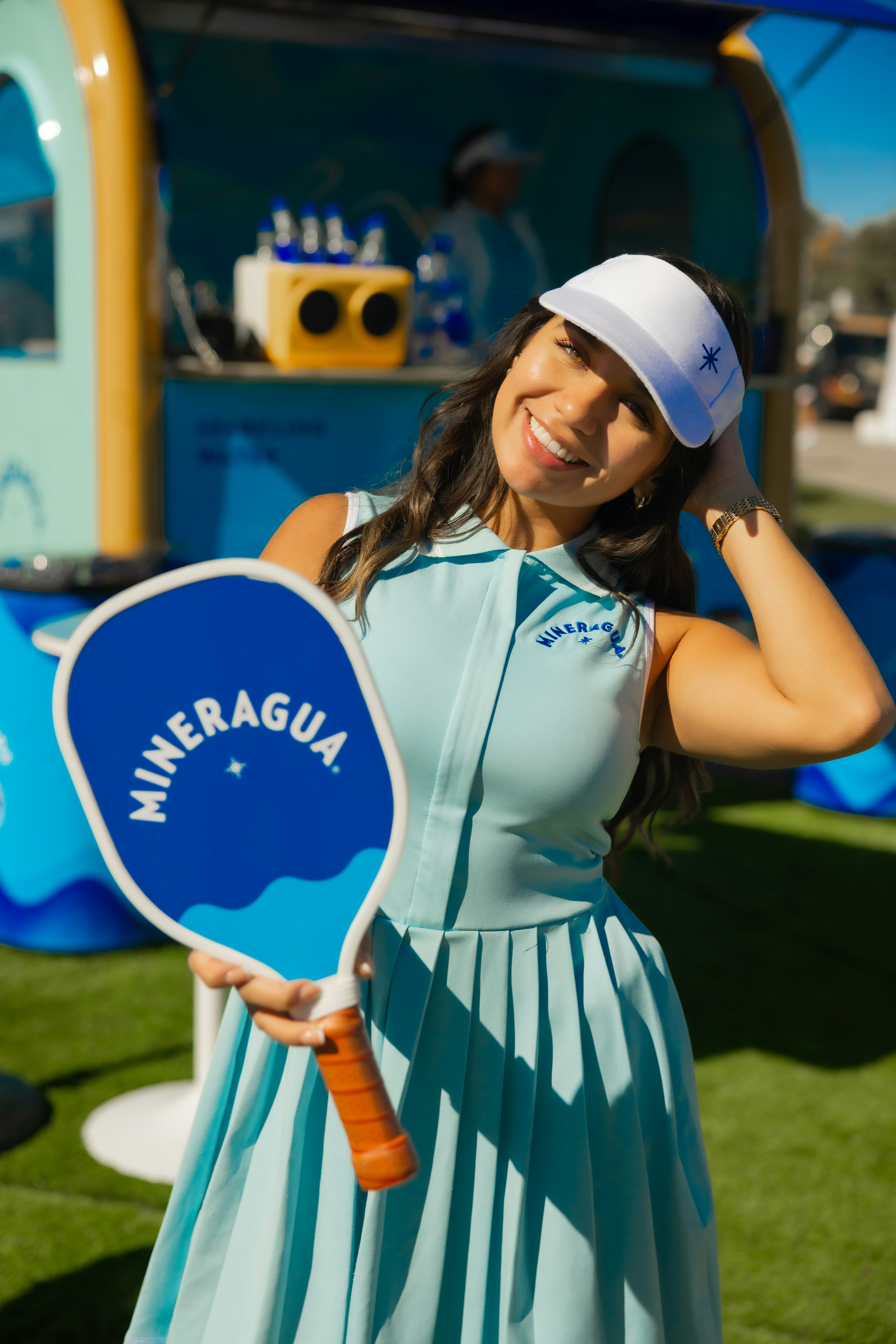 A smiling woman in a light blue dress holds a paddle with the Minaragua logo, set against a colorful beverage booth. Bright and inviting atmosphere.