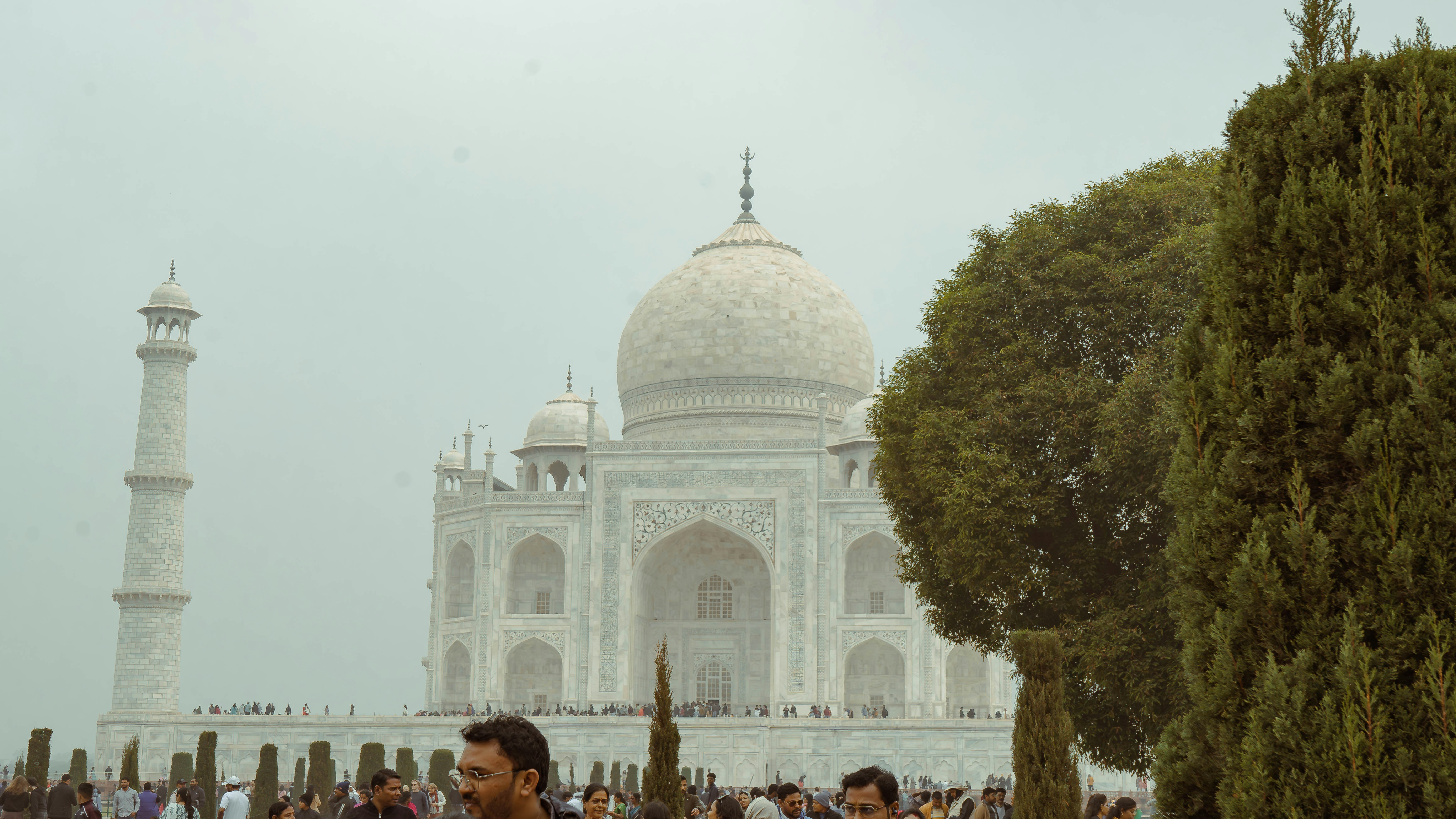 Taj Mahal surrounded by lush green trees under an overcast sky, with a crowd in the foreground.