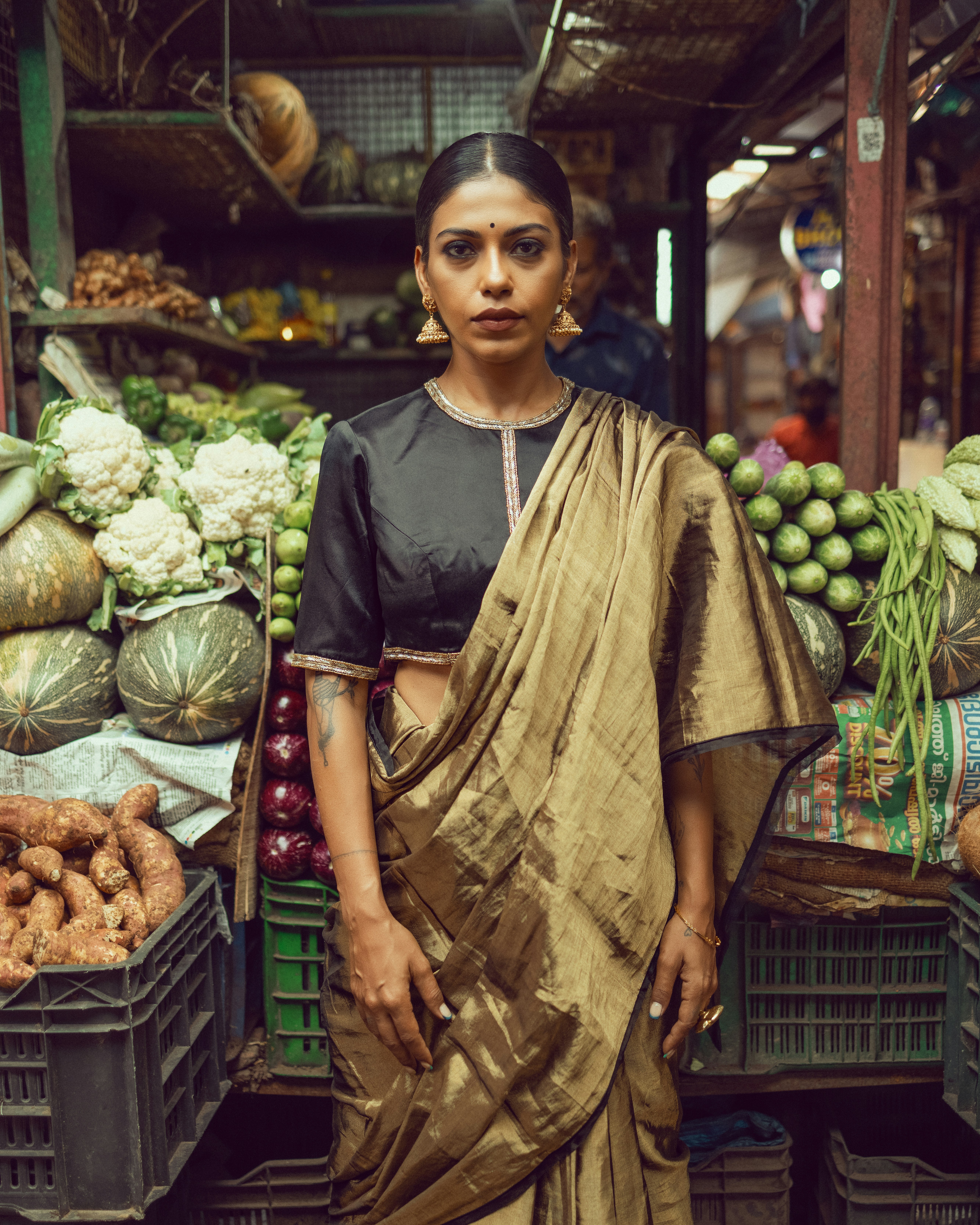 Woman in a gold and black sari stands amid colorful stacks of fresh vegetables in a lively market. Her attire's tones complement the vivid greens and whites of the produce.