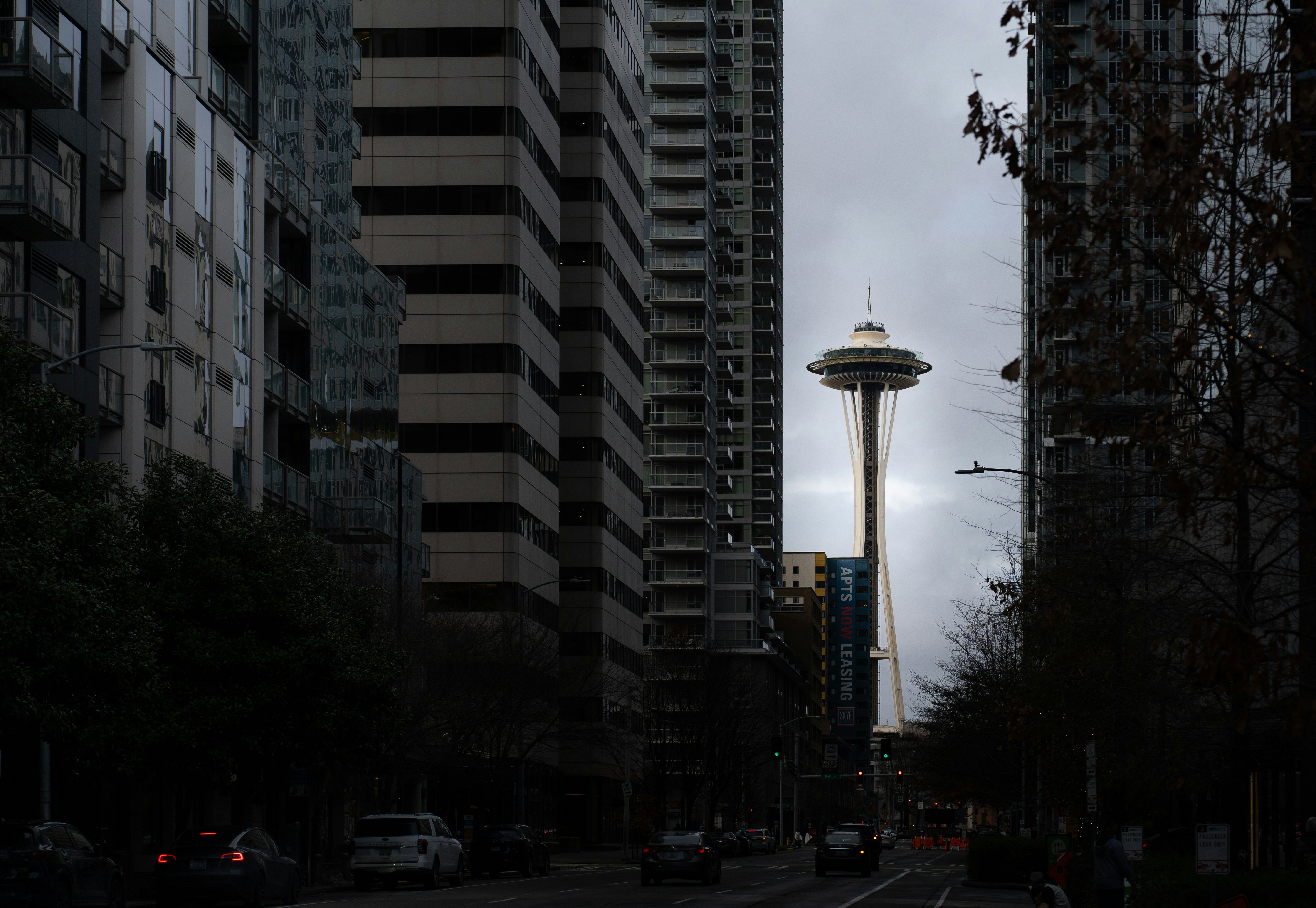 City street photo showing tall buildings lining a corridor toward the Space Needle silhouetted against a gray, overcast sky.