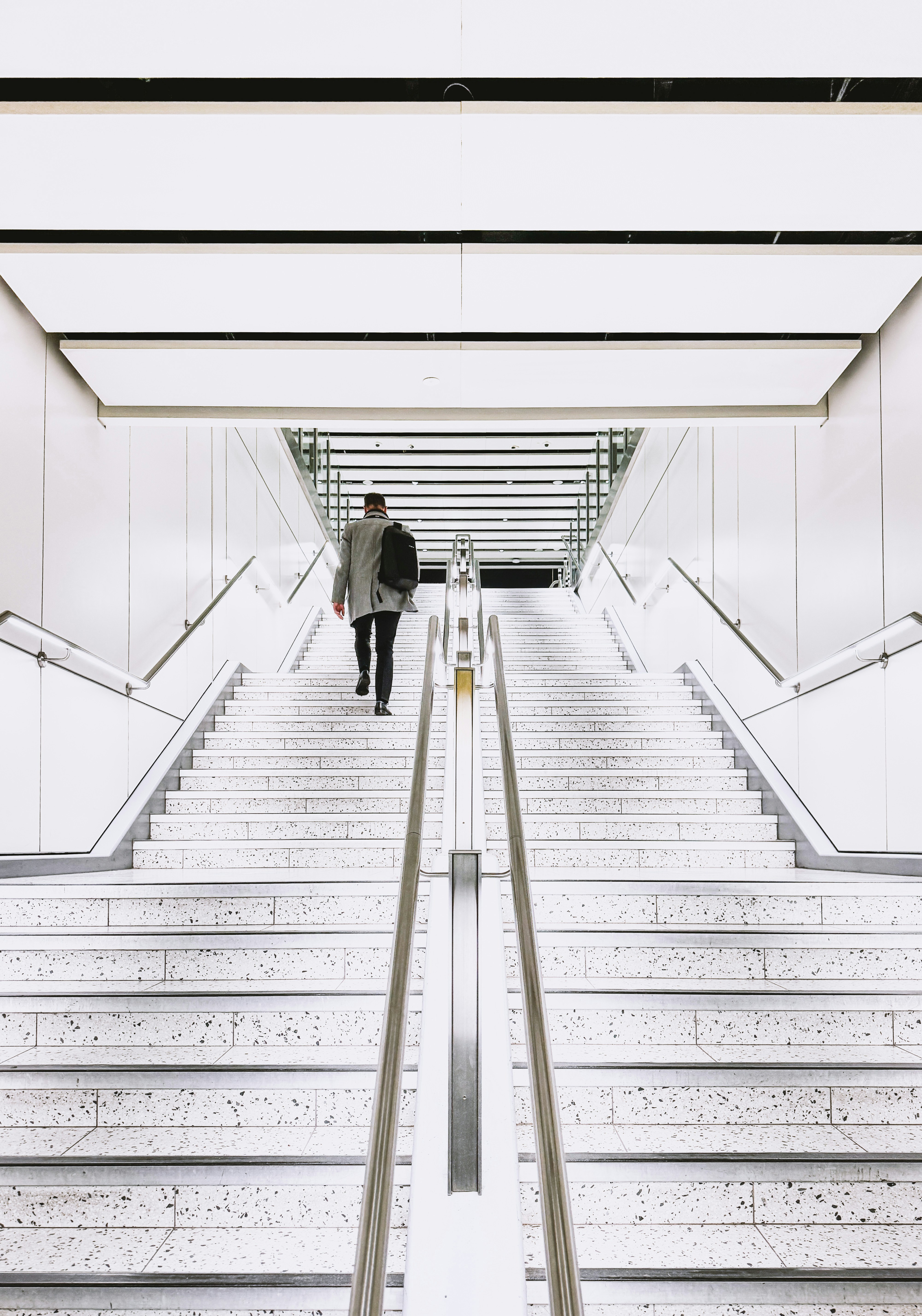 A lone figure navigates a bright, modern staircase surrounded by sleek white walls and contemporary lighting. The design emphasizes symmetry and open space.