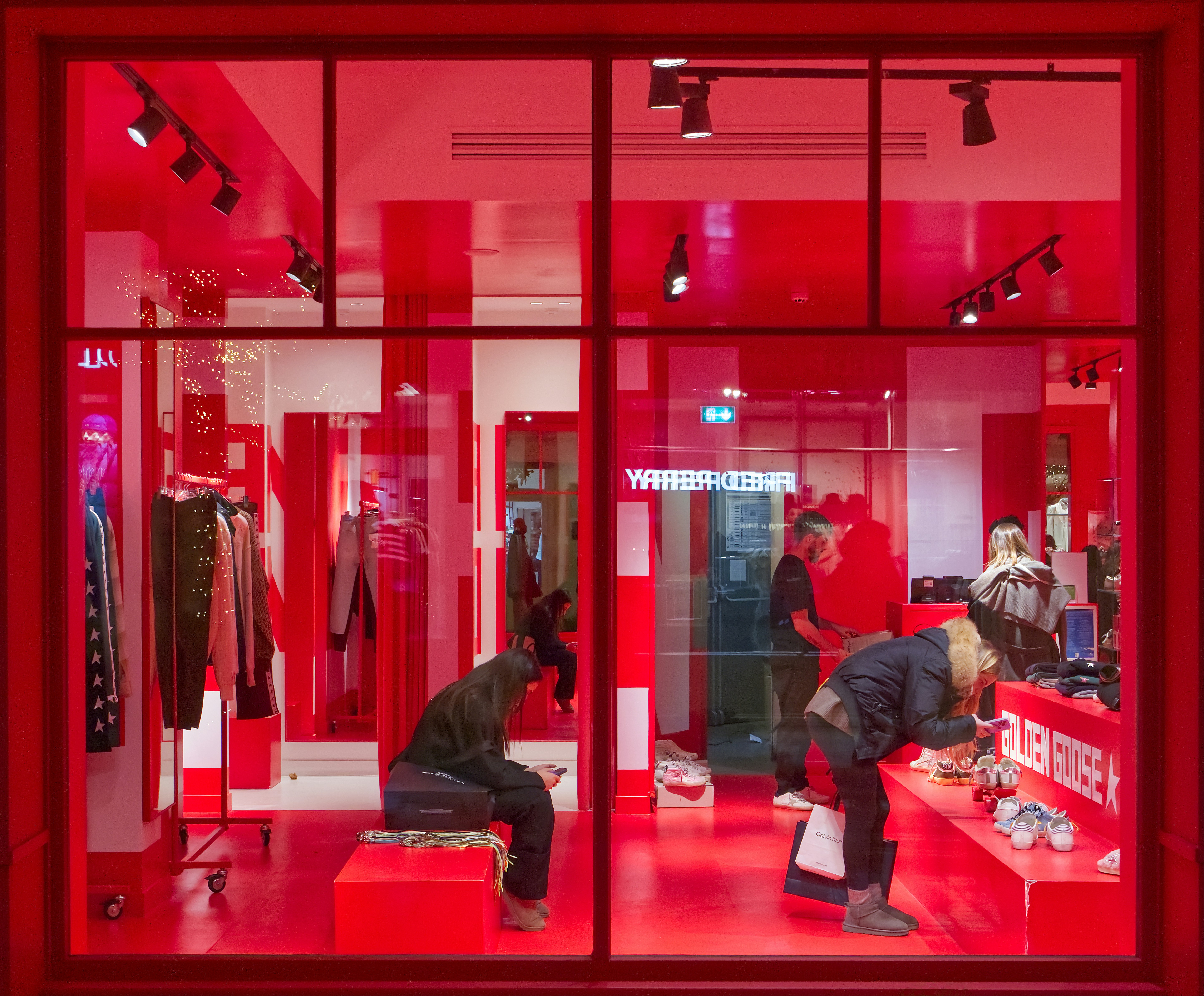 À travers l'écrin rouge flamboyant de cette vitrine, un théâtre urbain se dévoile, capturé dans une succession de cadres qui s'emboîtent tel un jeu de miroirs ou une composition de type Mondrian. La devanture du magasin se transforme en une composition graphique, où chaque rectangle vitré révèle un fragment de cette scène de shopping contemporaine.