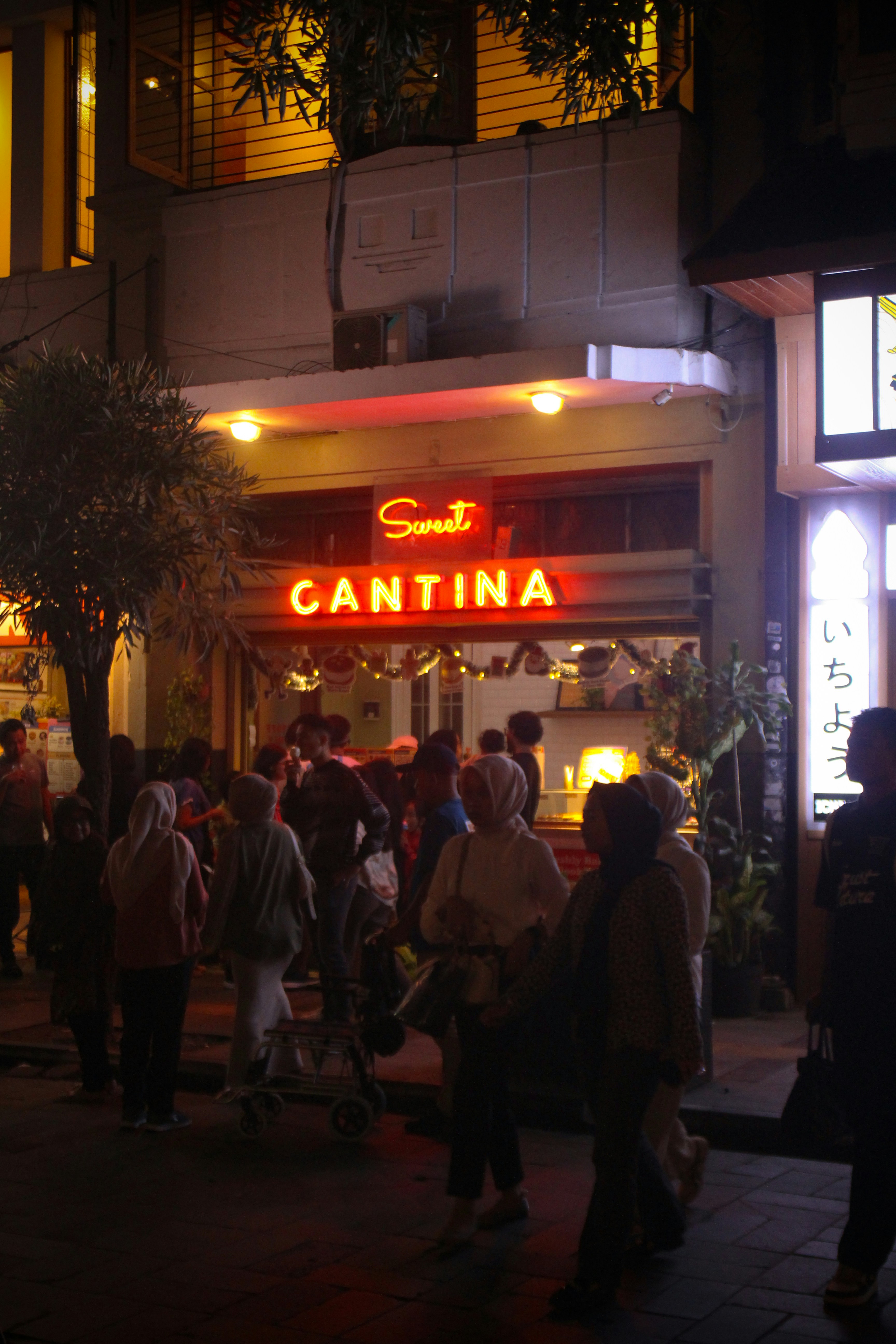 Neon-lit cantina storefront at night draws a crowd along the sidewalk, with warm red signage dominating the scene.
