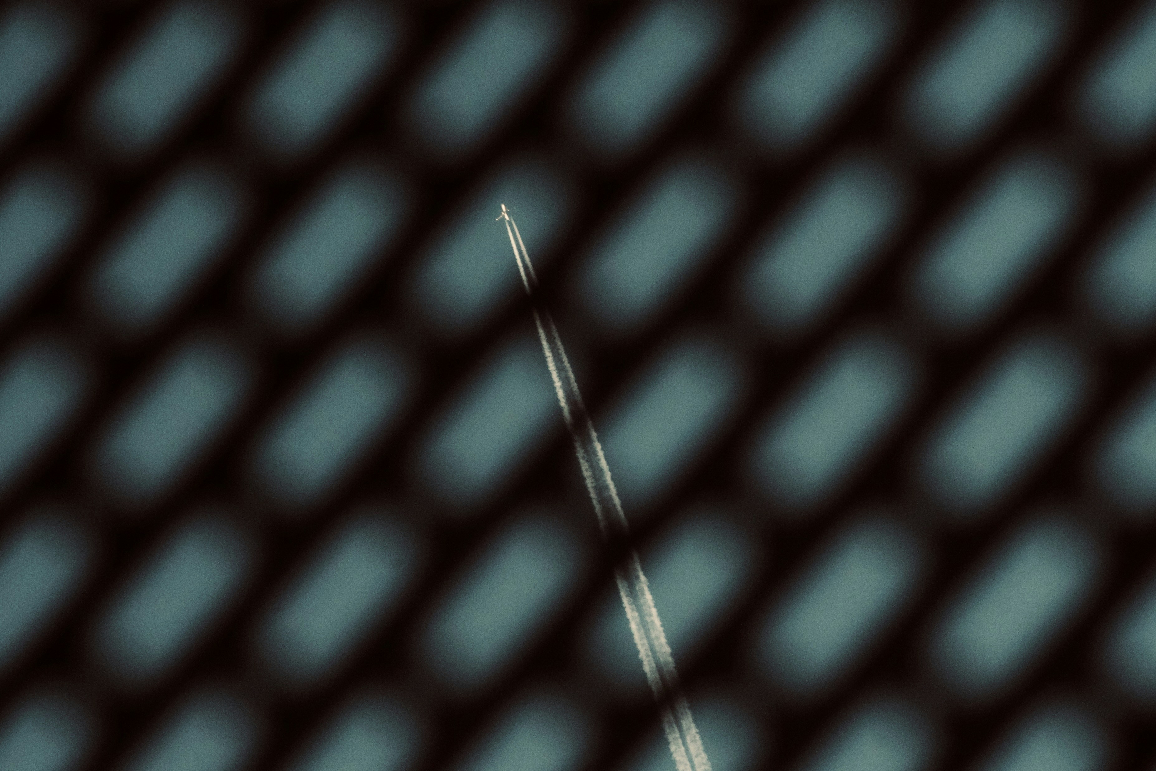 Airplane leaving a trail of contrails against a textured backdrop of a fence.