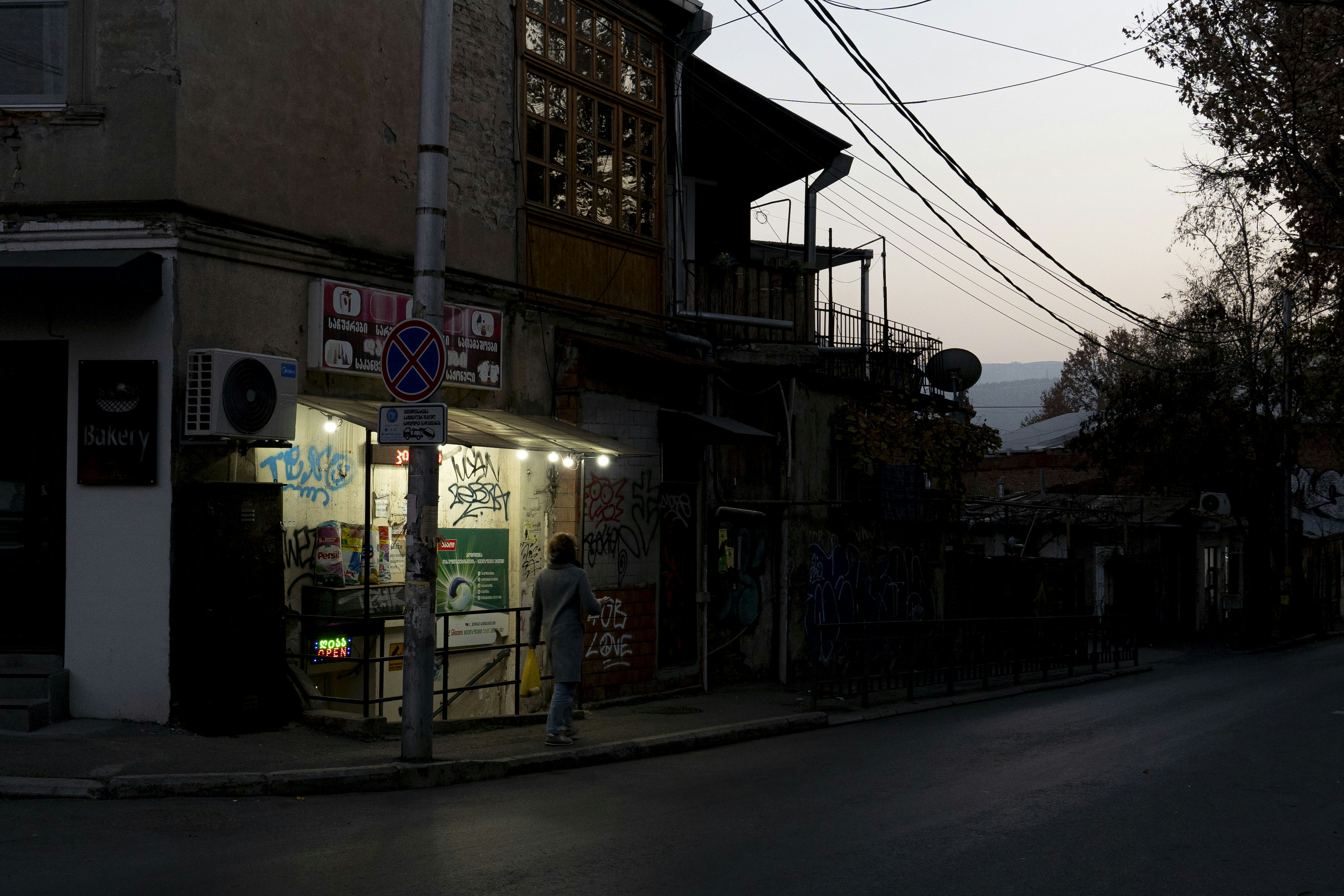 Dimly lit street corner with a small shop illuminated at dusk.